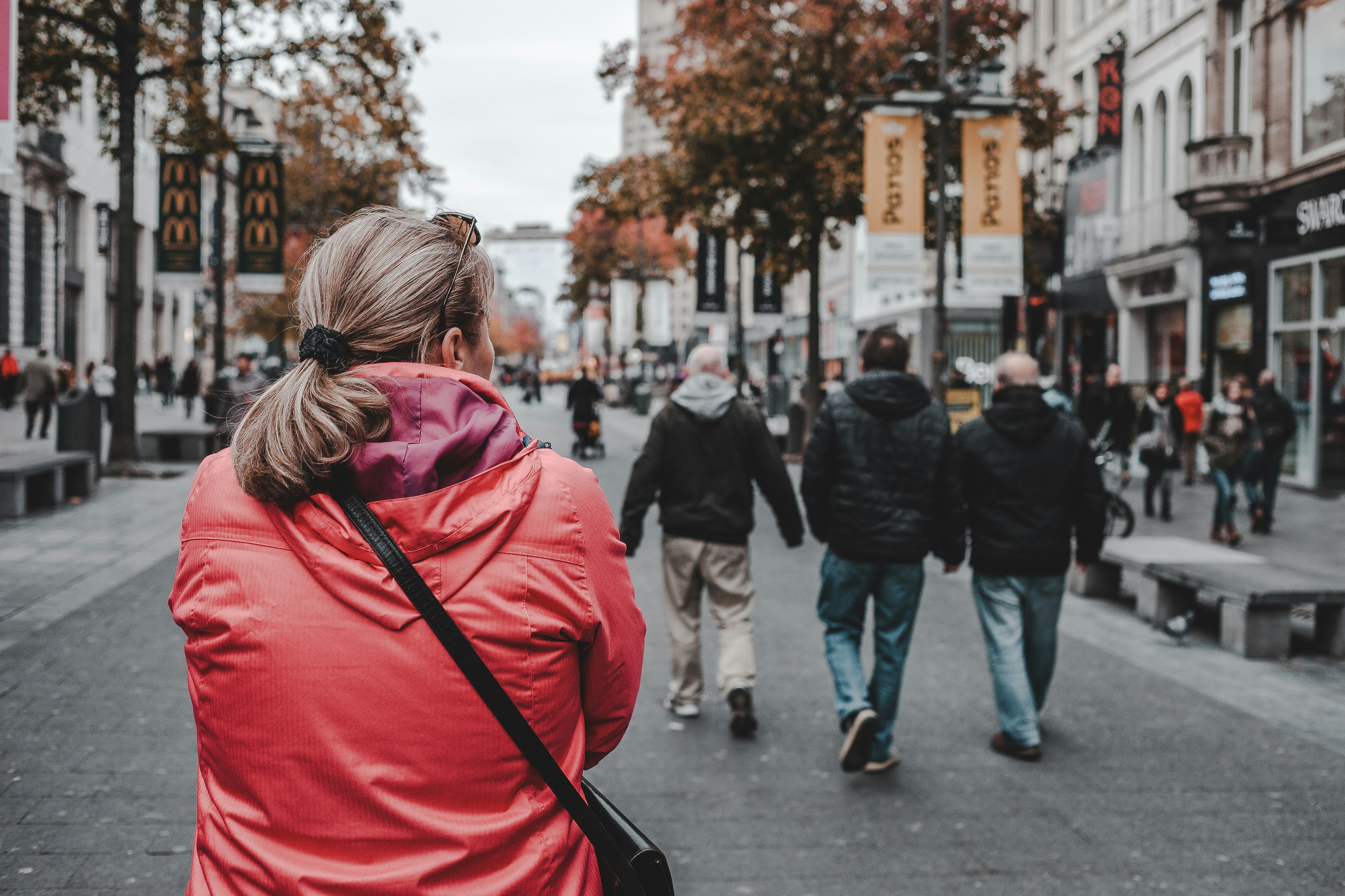 Person in a red jacket walking along a busy Antwerp street lined with autumn trees.