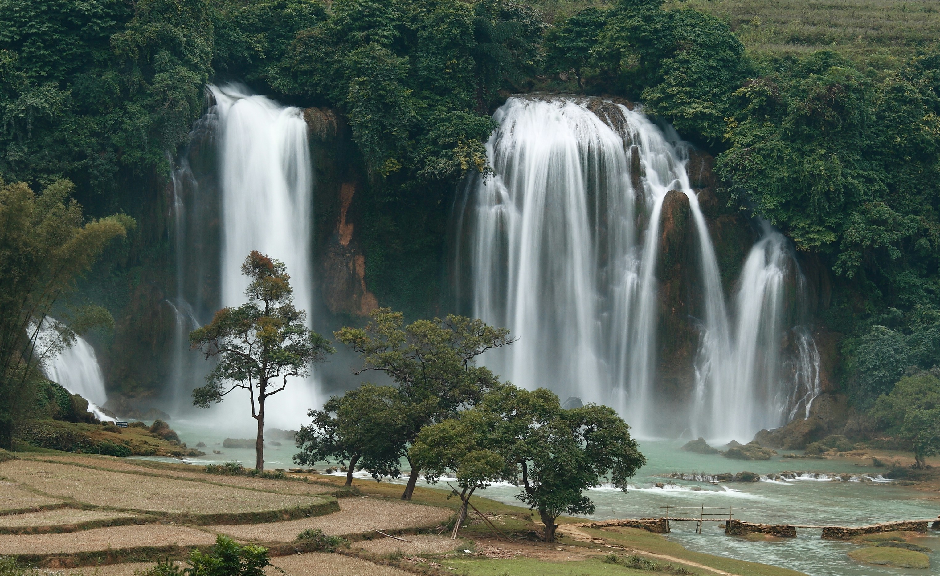 Majestic waterfalls cascade over lush green cliffs into a serene pool surrounded by trees and terraced fields.