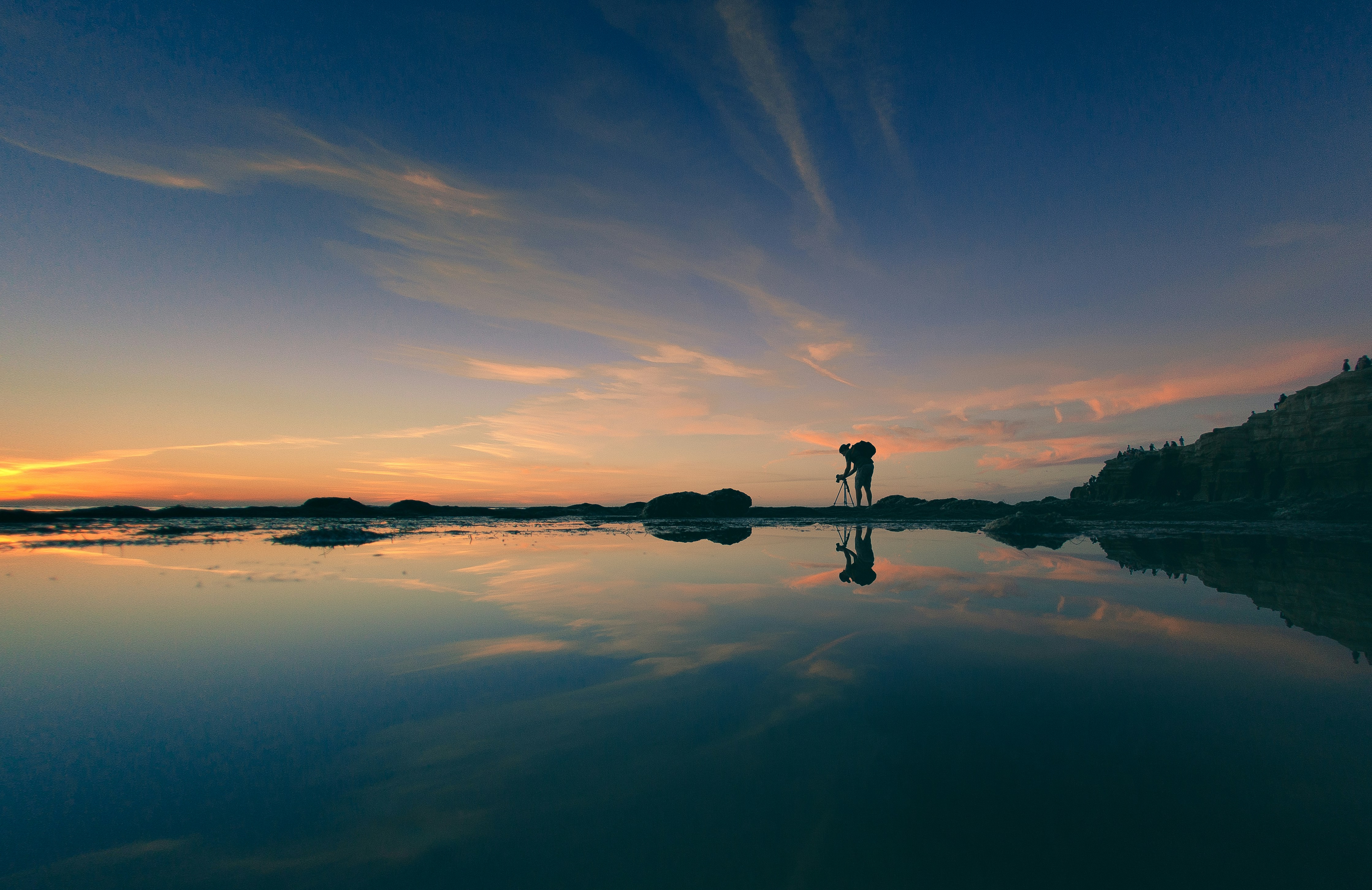 Silhouette of a person adjusting a camera tripod against a vibrant sunset, reflected in calm tidal waters.