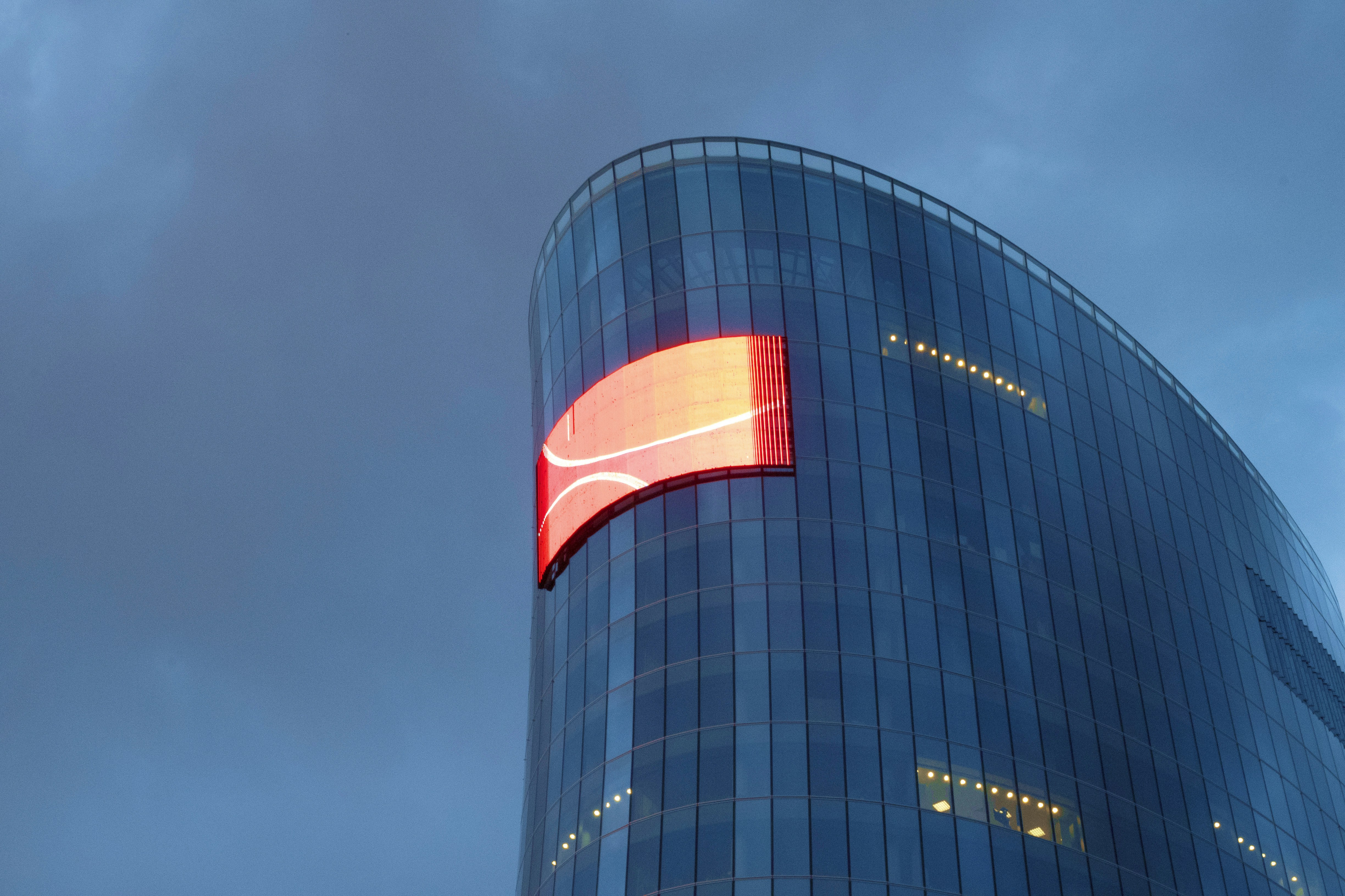 Curved glass skyscraper with a vibrant red digital display against a moody evening sky.