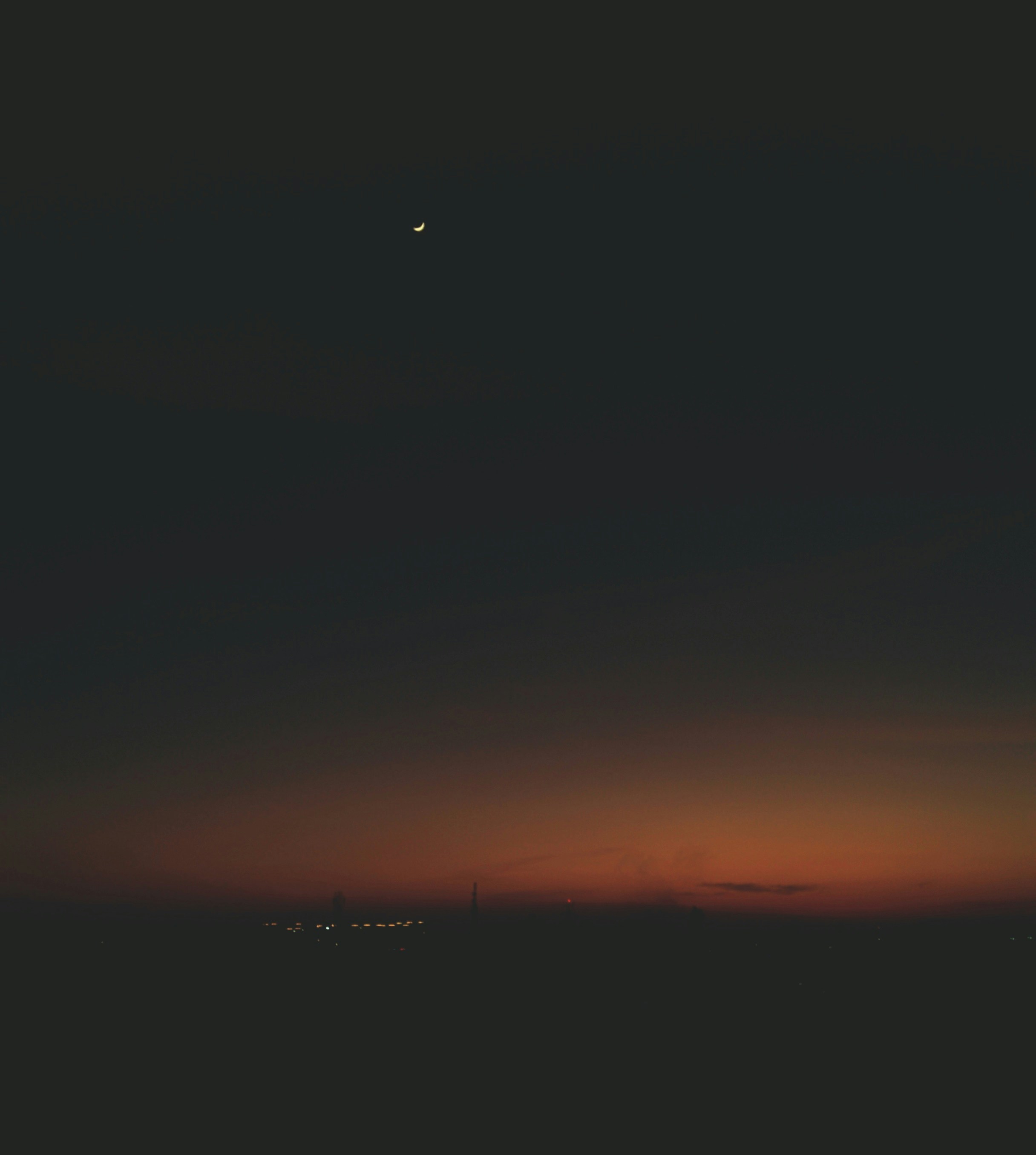 A slender crescent moon glows above a quiet horizon, where a warm orange glow fades into night.