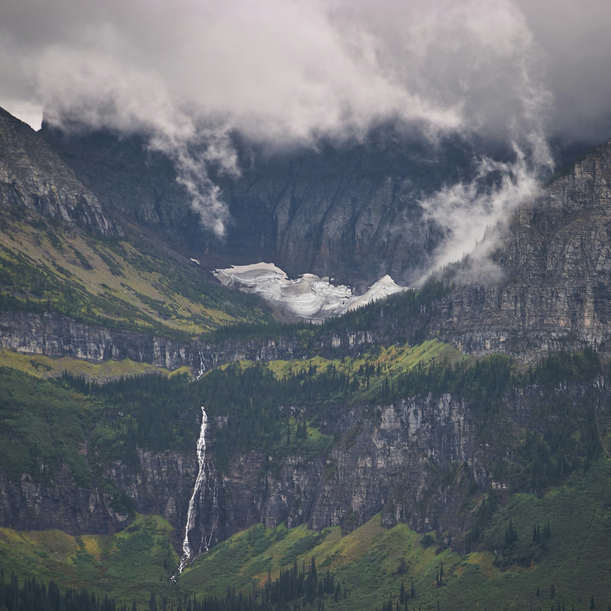 A dramatic waterfall cascades down rugged cliffs, revealing a glimpse of a glacier above, shrouded in mist and clouds.