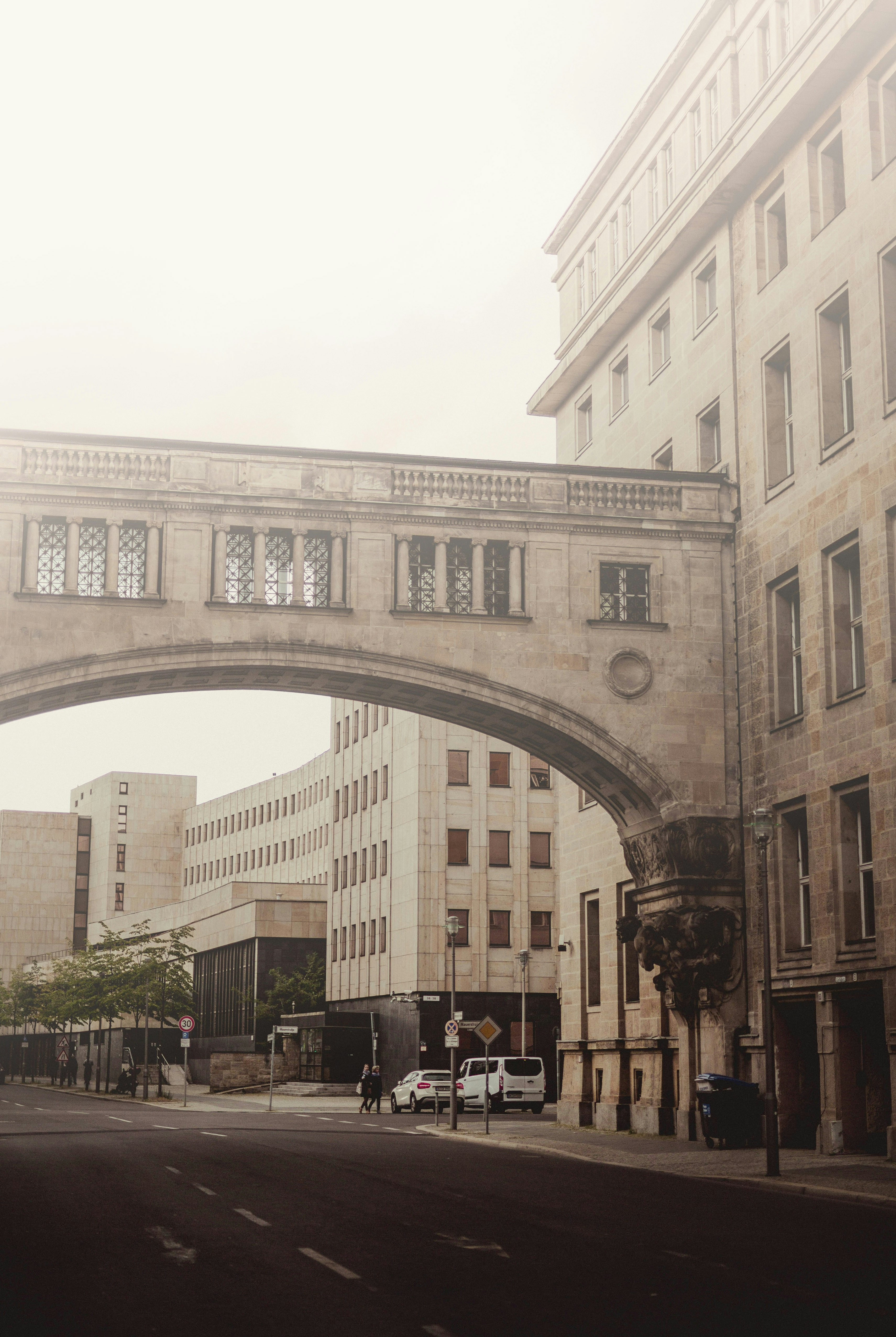 An ornate stone bridge connecting two buildings in a modern urban setting, showcasing a blend of historical and contemporary architecture.