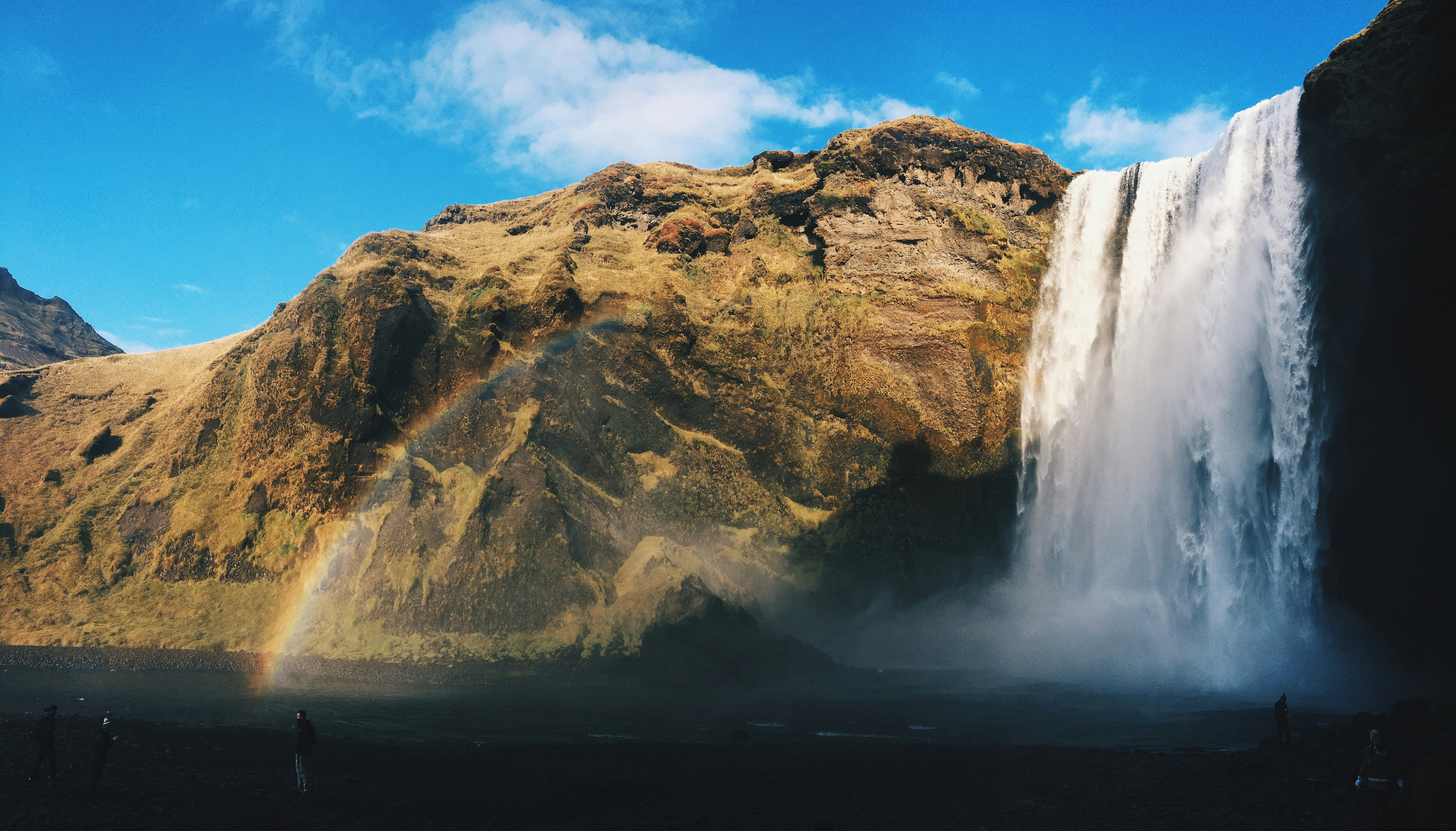 Majestic waterfall flowing beside a vibrant rainbow arcing over rugged cliffs under a clear blue sky.
