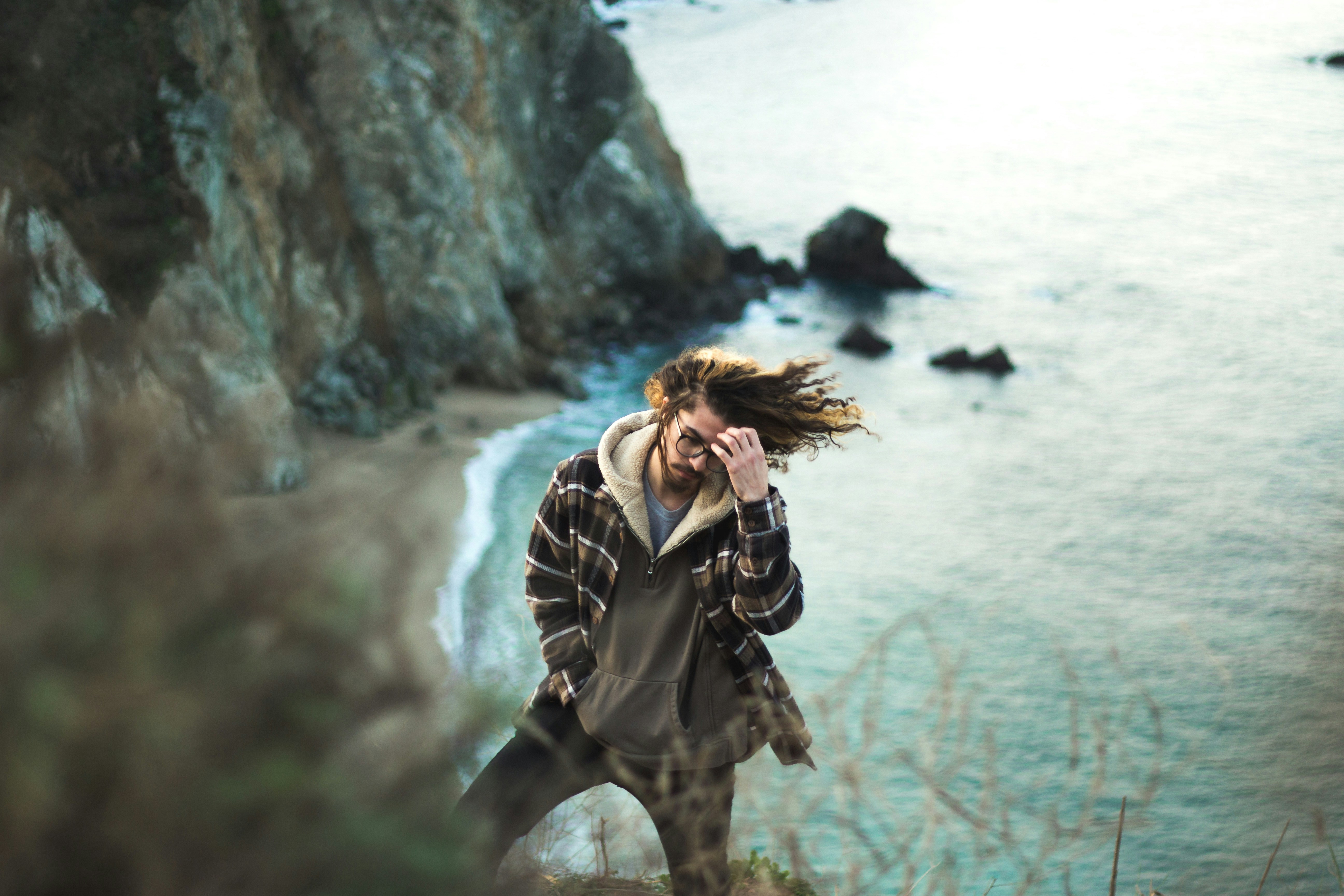 Individual standing on a cliff, hair flowing in the wind, overlooking a serene coastline. The rugged landscape contrasts with the calm ocean below.