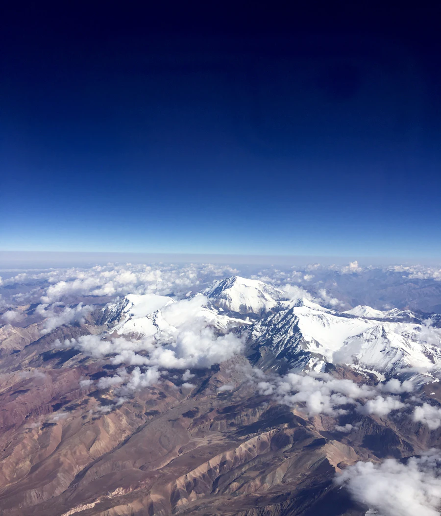 Aerial view of a snow-covered mountain range rising above scattered clouds under a deep blue sky. Rugged brown ridges stretch across the foreground.