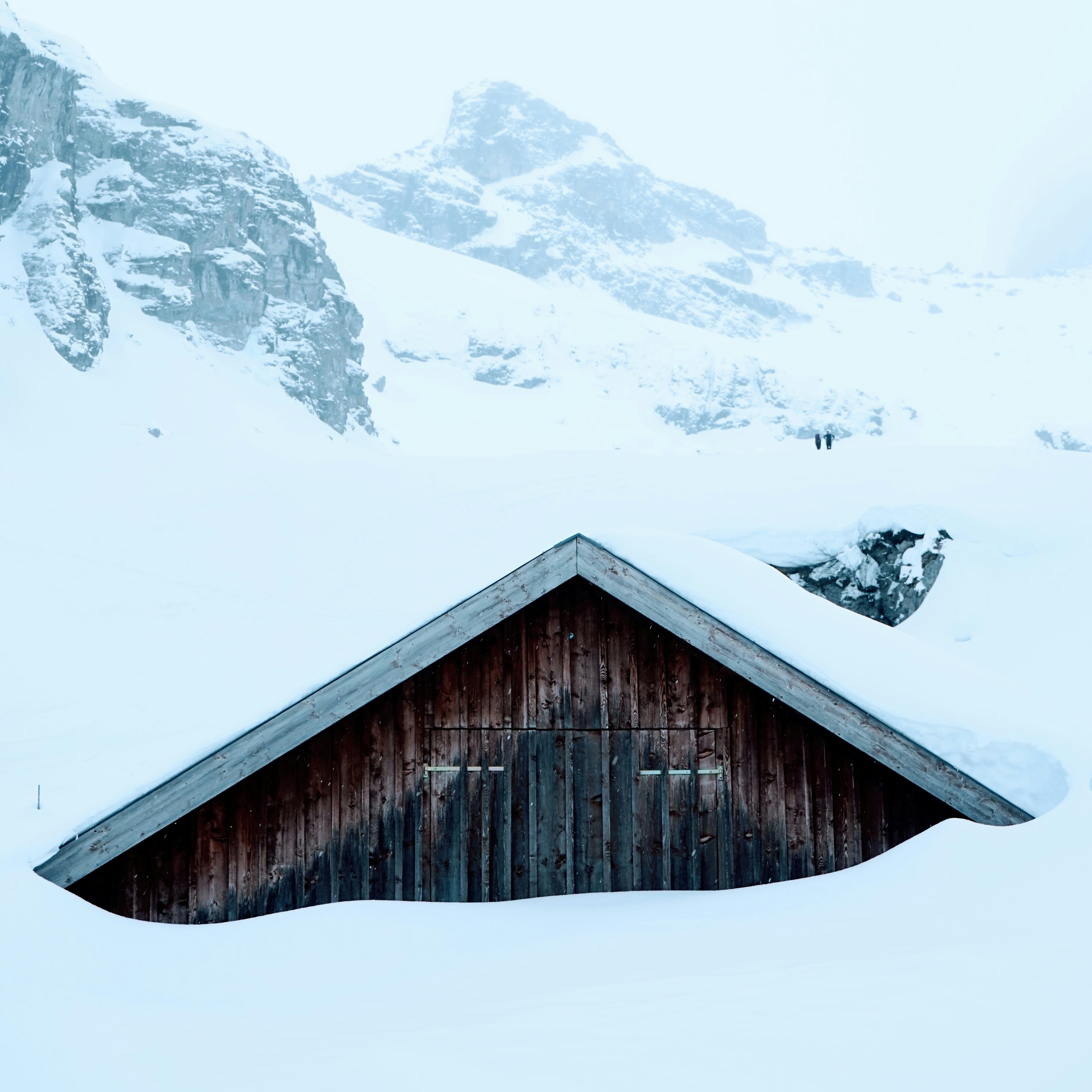 Wooden cabin roof peeks through deep snow against a backdrop of rugged, snow-covered mountains.