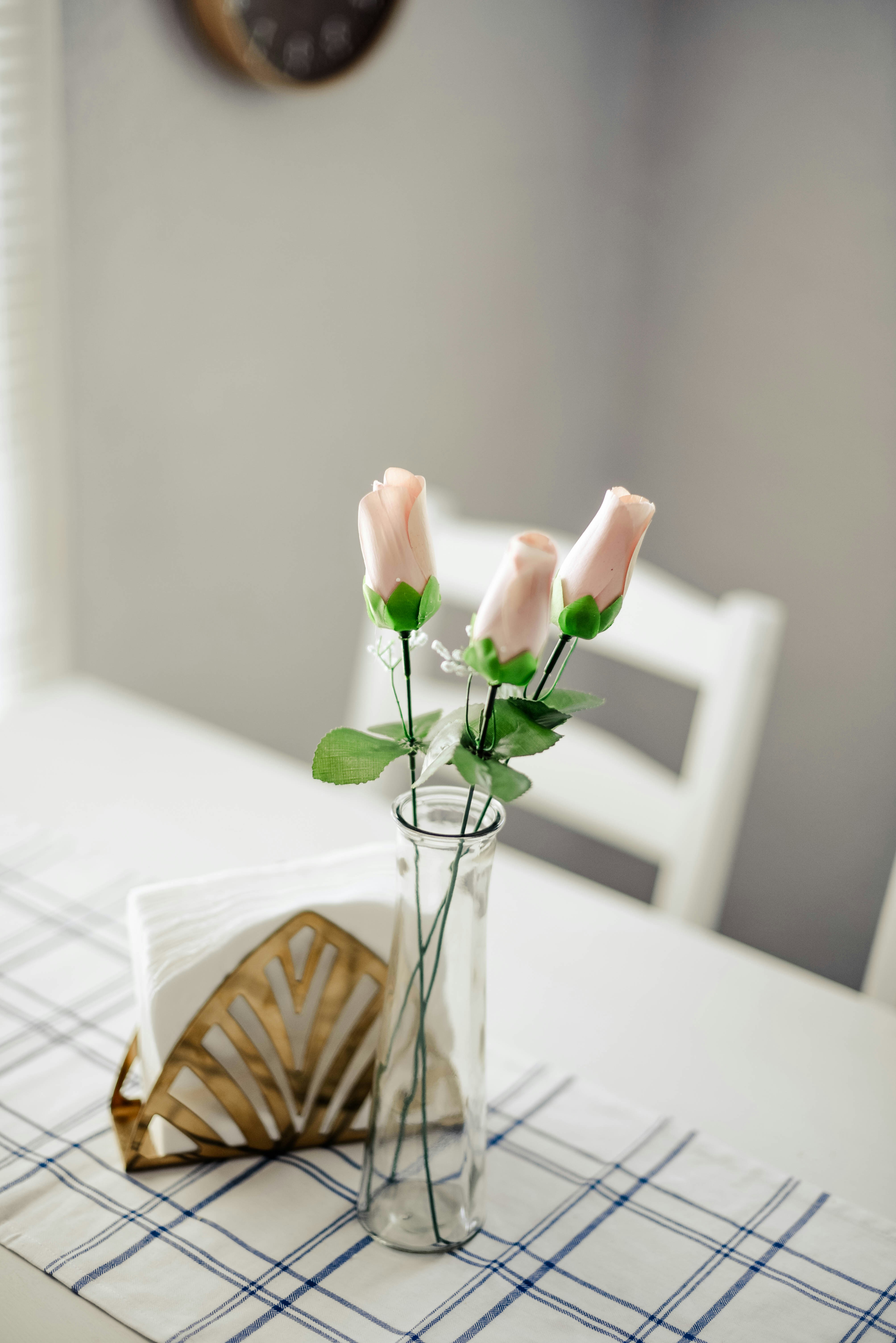 Three delicate pink roses in a slender glass vase, accompanied by a stylish napkin holder on a checkered tablecloth.