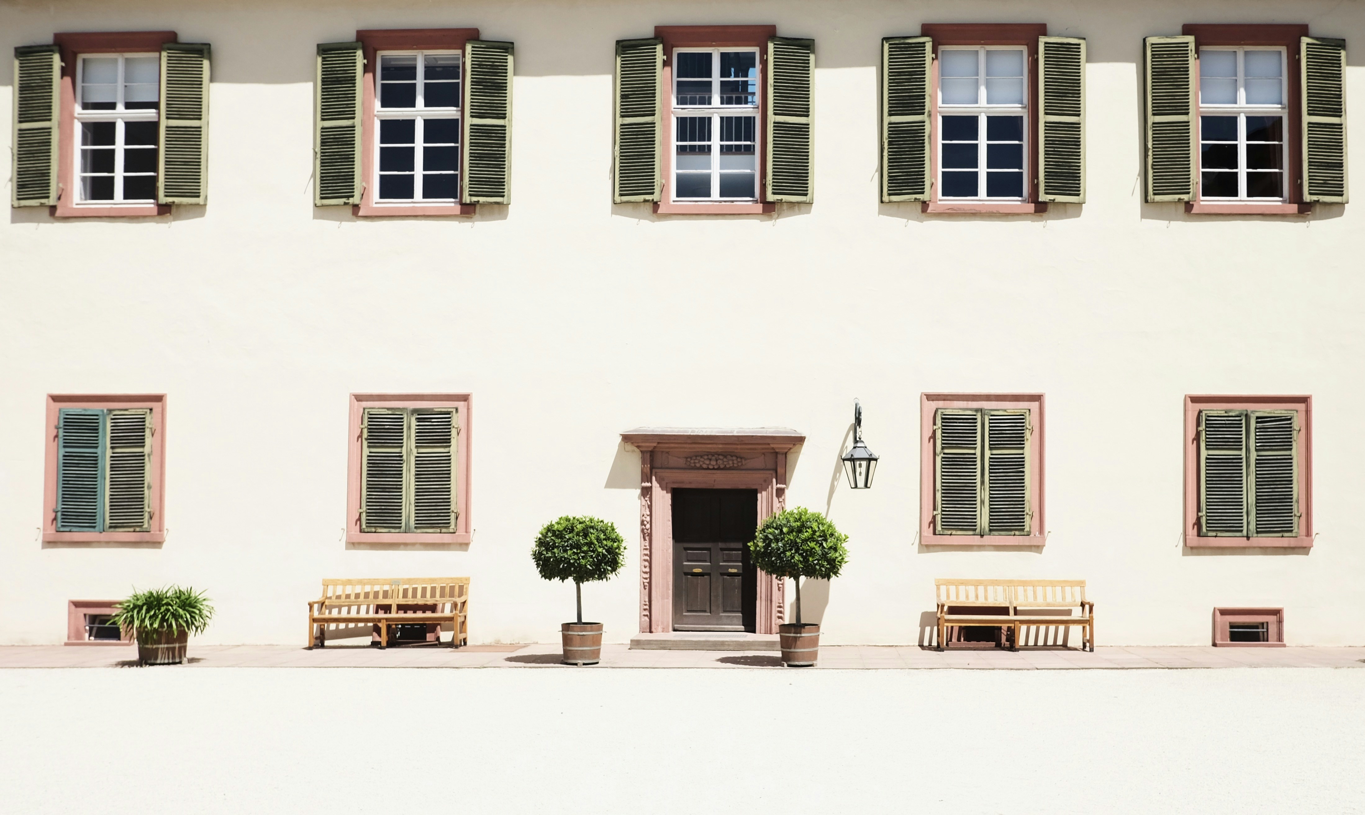 Facade of a building with evenly spaced windows and green shutters, flanked by benches and potted trees.