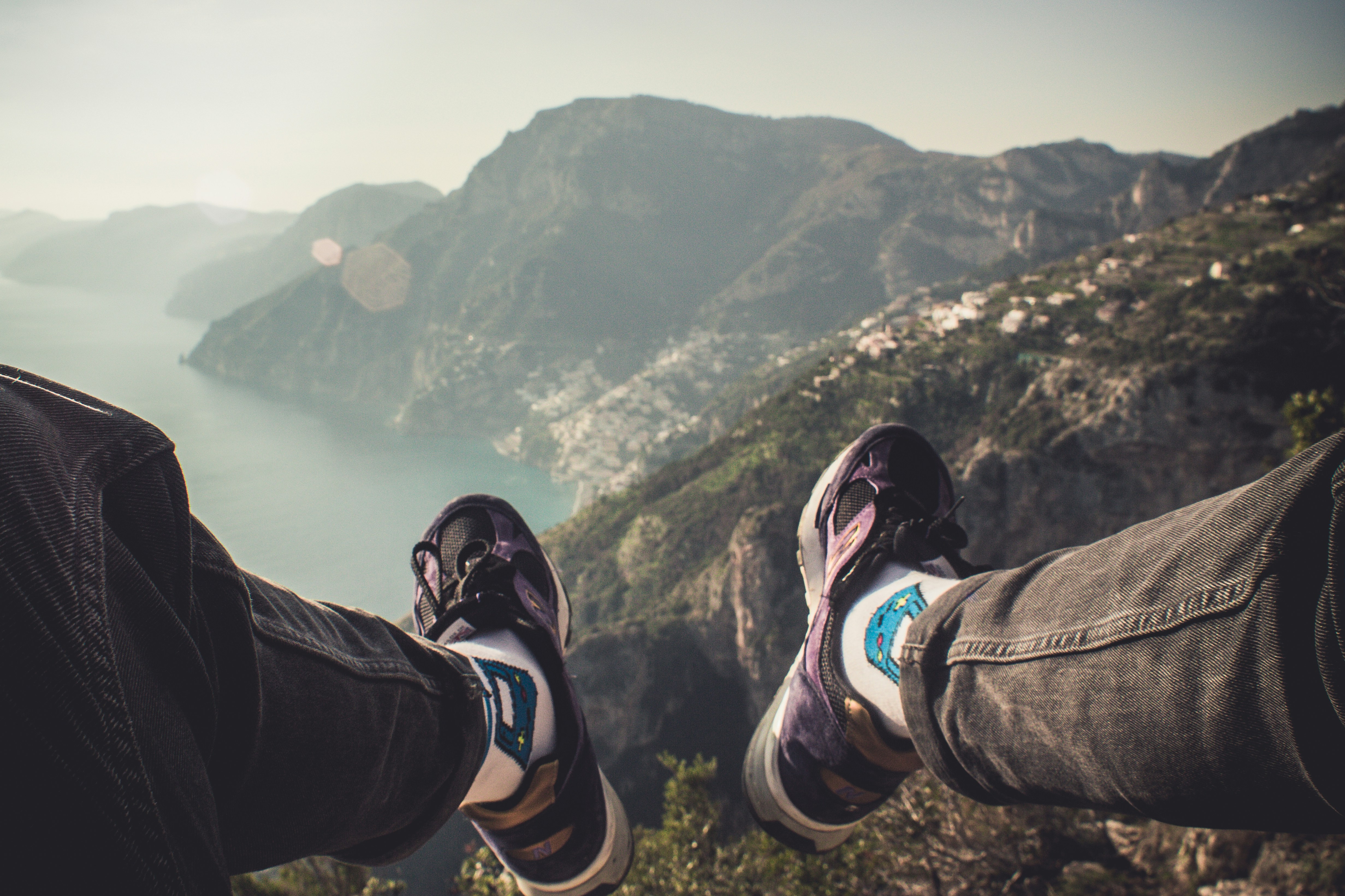 Feet in colorful sneakers dangle over a high cliff with a vast coastal landscape in the background.