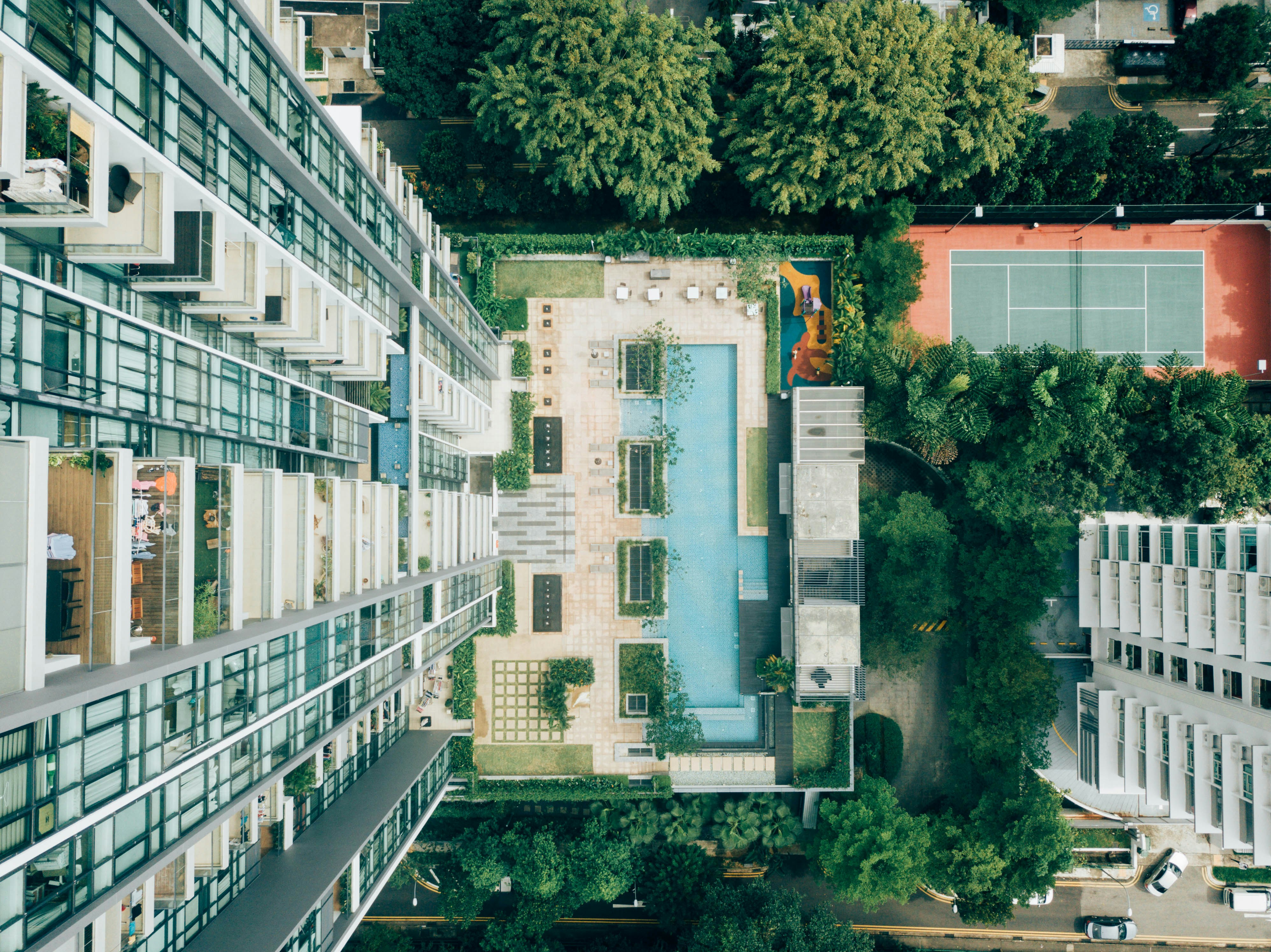 This aerial image captures a stunning top-down view of a modern residential complex with a central swimming pool and surrounding greenery. The composition is enhanced by the geometric lines of the buildings, the vibrant blue of the pool, and the lush green foliage, creating a striking contrast against the urban backdrop. The lighting is soft, casting gentle shadows that add depth and texture to the scene, making it both visually captivating and serene.