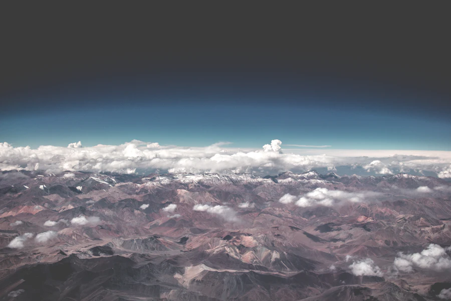 Aerial landscape photograph of layered mountain ridges beneath a gradient blue sky with wispy clouds along the horizon. The composition emphasizes vast elevation and distant terrain.