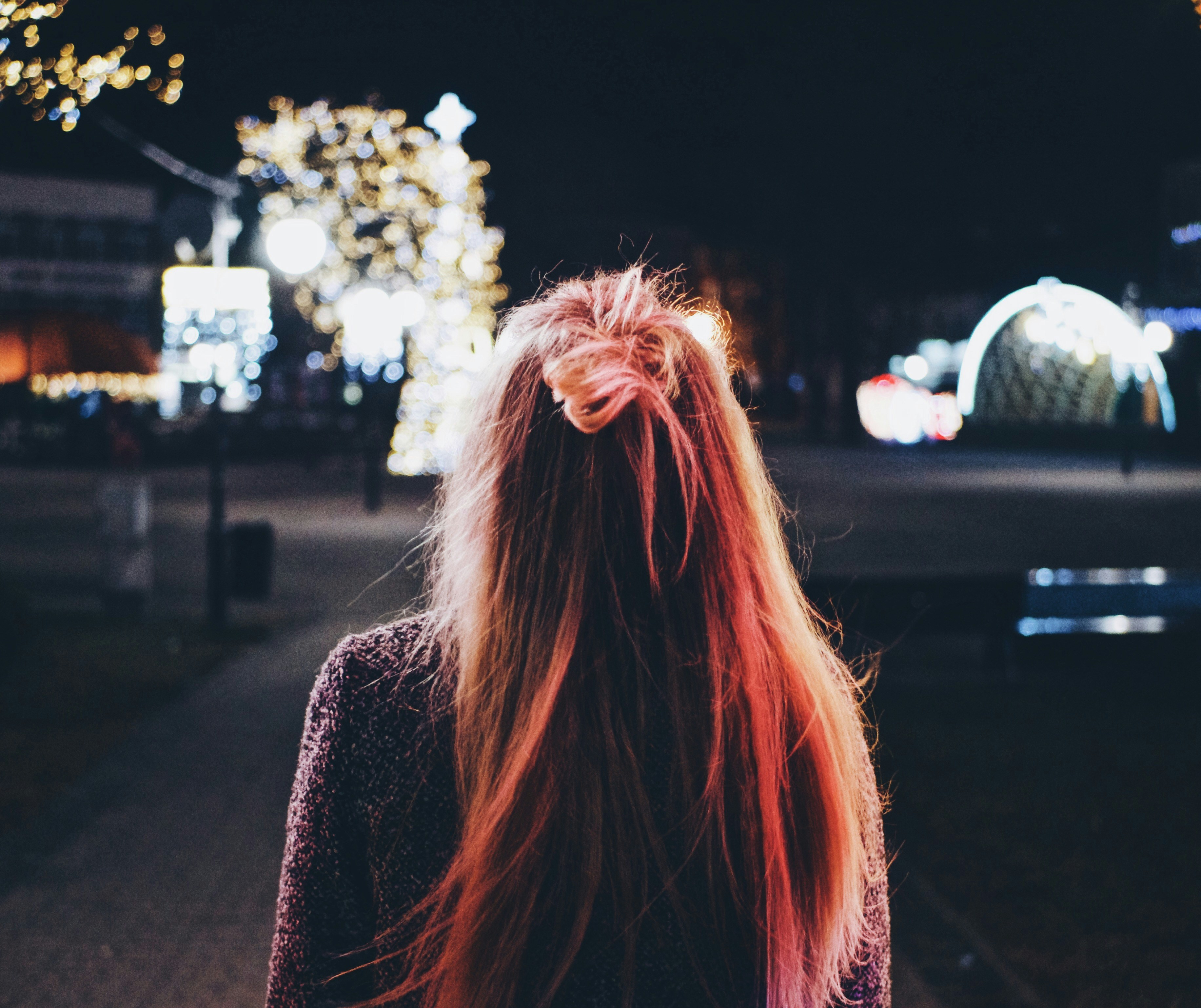 Person with long hair standing in a dimly lit street with blurred festive lights.