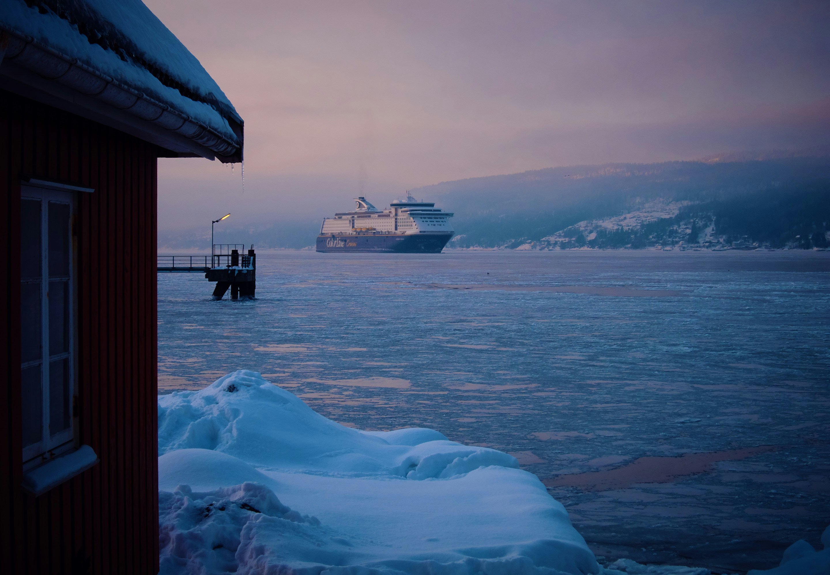 A large cruise ship navigating through icy waters, framed by a rustic red cabin and a snow-covered landscape at dawn.