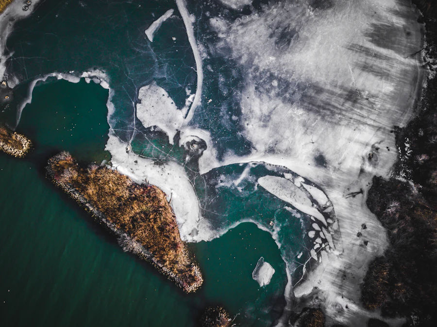 Aerial view of a frozen shoreline with dark teal water, white ice sheets, and a narrow rocky peninsula. Wind-swept snow streaks texture the upper right edge.