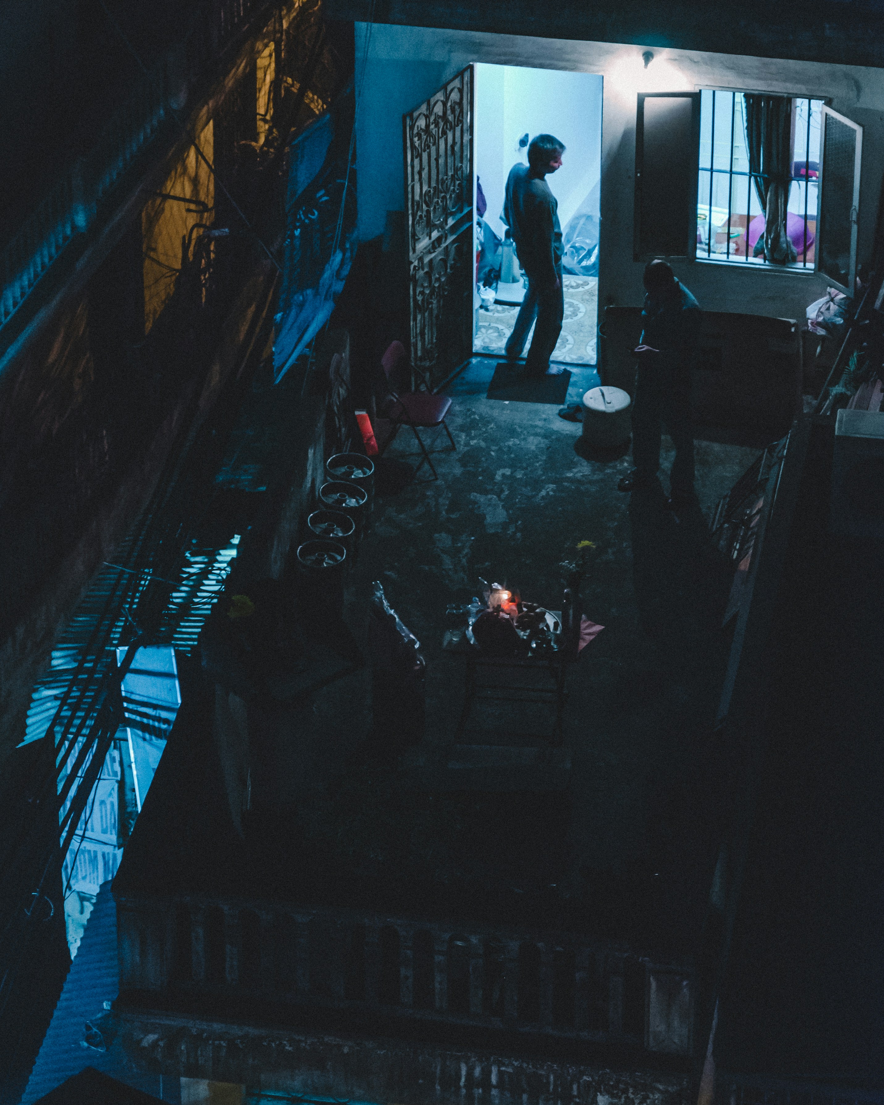 Dimly lit rooftop scene with figures engaged in quiet activities, framed by open doors and contrasting shadows. A glimpse into urban life after dark.