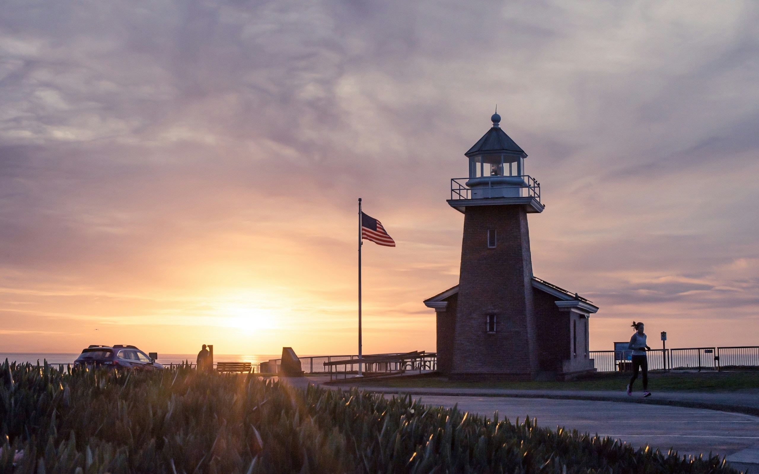 Jogger passing a lighthouse and flagpole against a vibrant sunset sky.