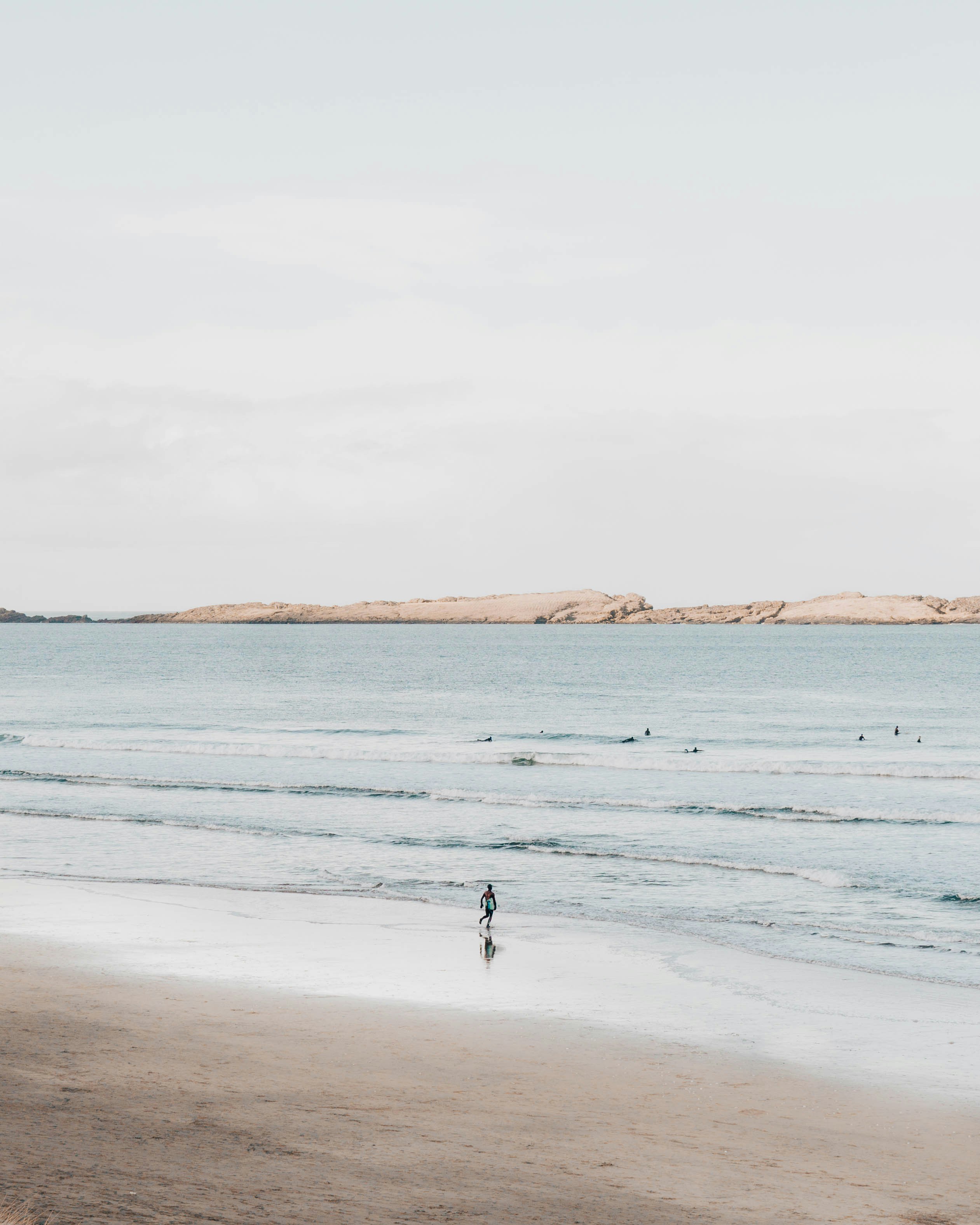 A lone figure walking along the wet sand of a tranquil beach, with gentle waves lapping at the shore under a soft, cloudy sky.