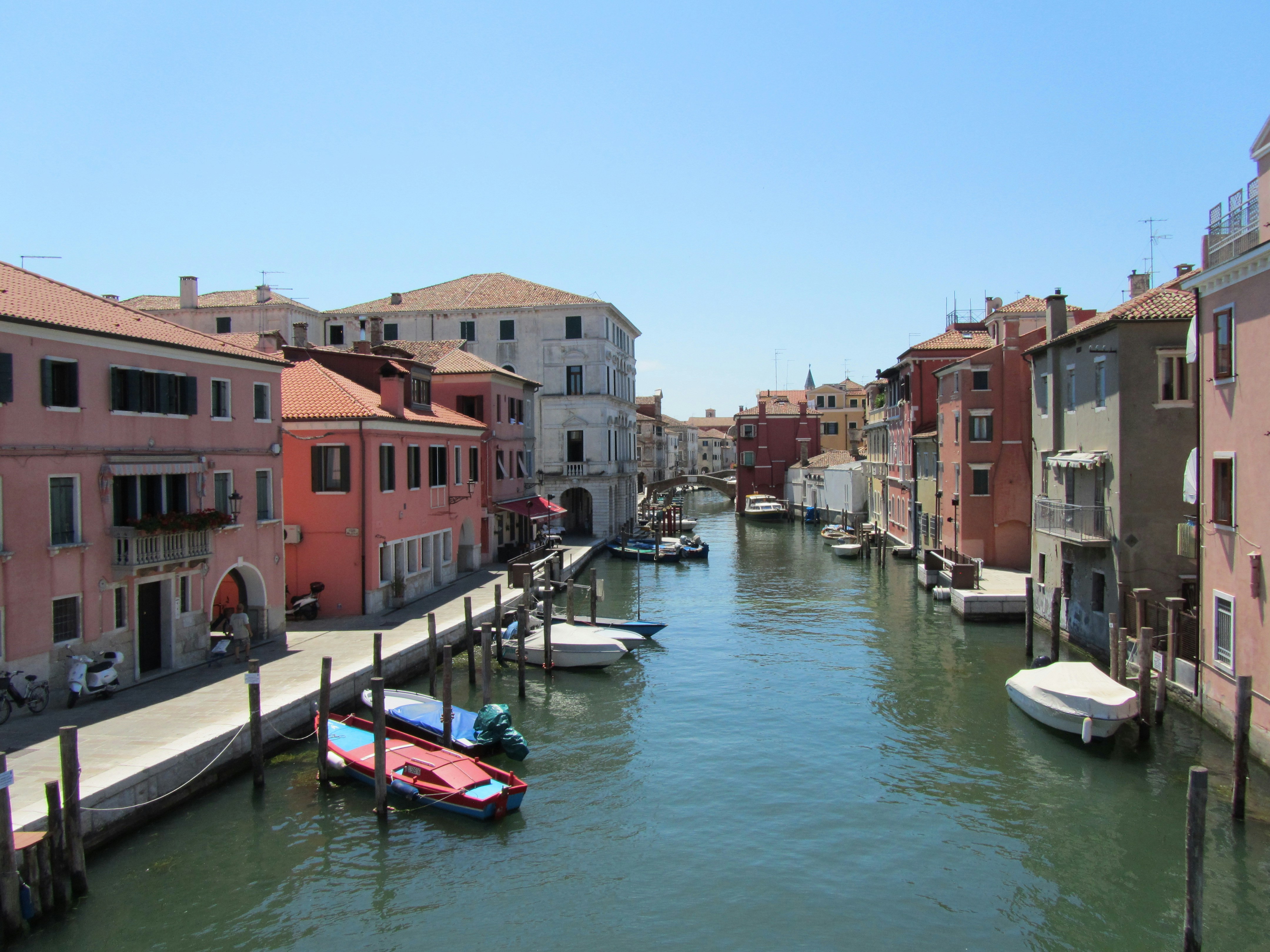 Serene canal flanked by pastel-hued buildings with boats moored along the edges under a clear blue sky.