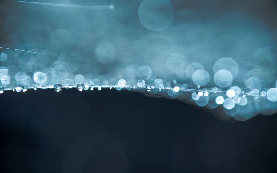 Soft blue abstract scene with a dark curved foreground edge lined by tiny bright droplets and out-of-focus light circles above it.