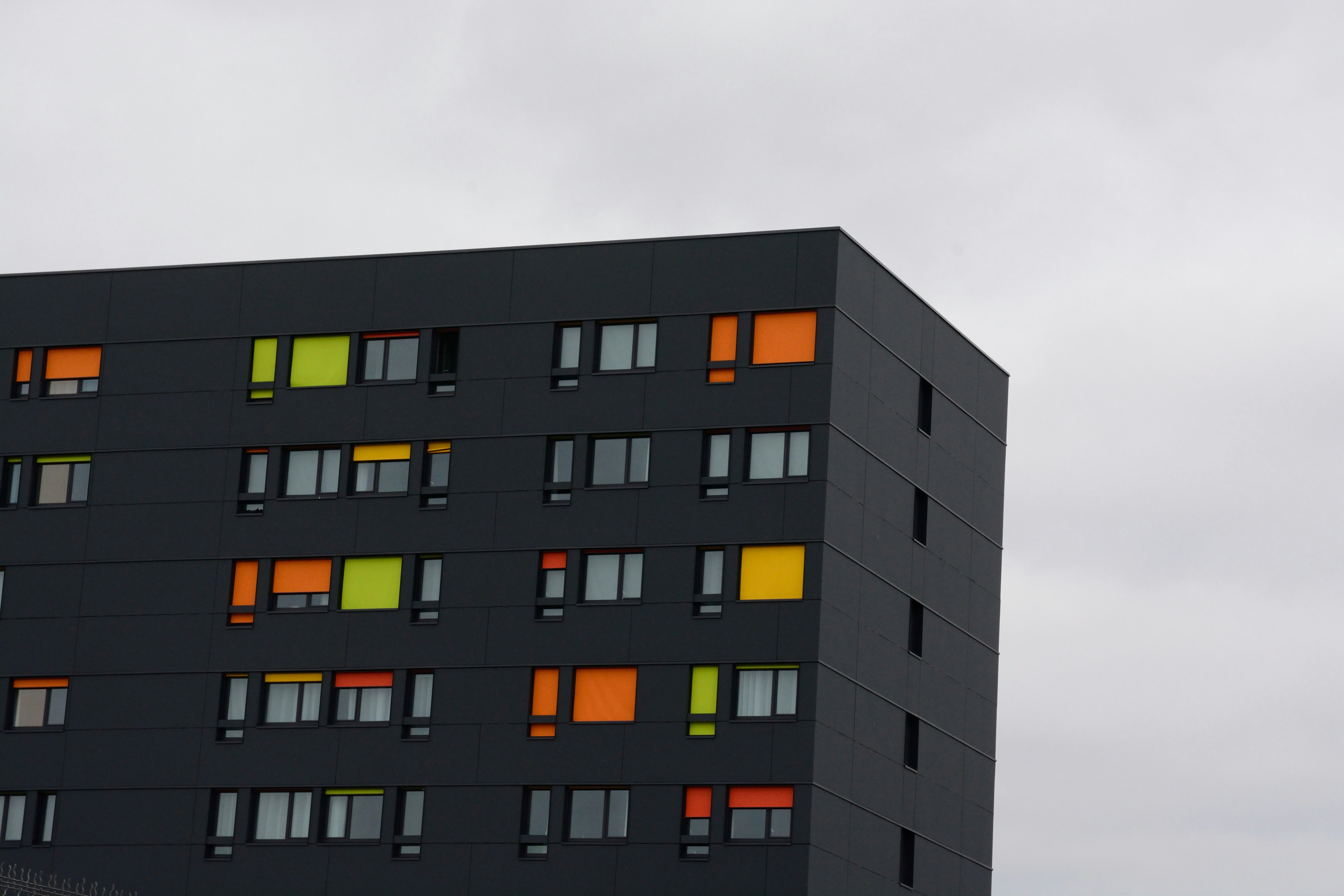 Modern building facade featuring a grid of colorful windows in orange and green against a dark exterior. The cloudy sky adds a dramatic backdrop.