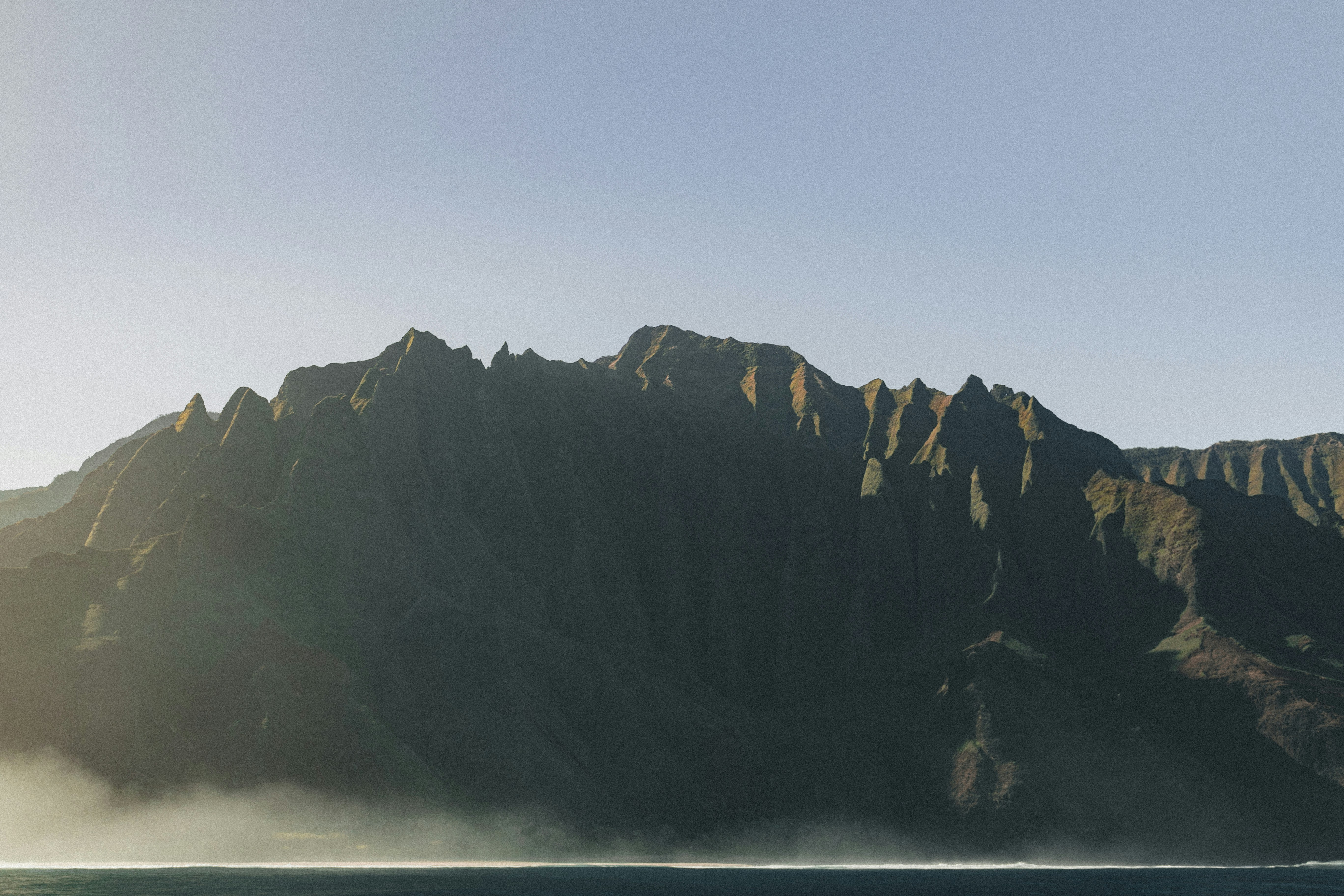 Dramatic mountain range of the Na Pali Coast silhouetted against a clear sky, with soft mist rising from the ocean below.