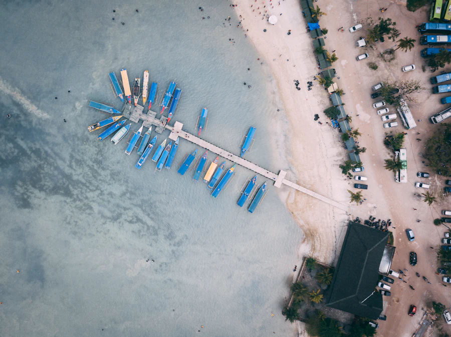 Top-down view of a narrow pier extending into pale water, with blue boats moored along both sides. A sandy beach, parked cars, and a dark-roofed building frame the shoreline.