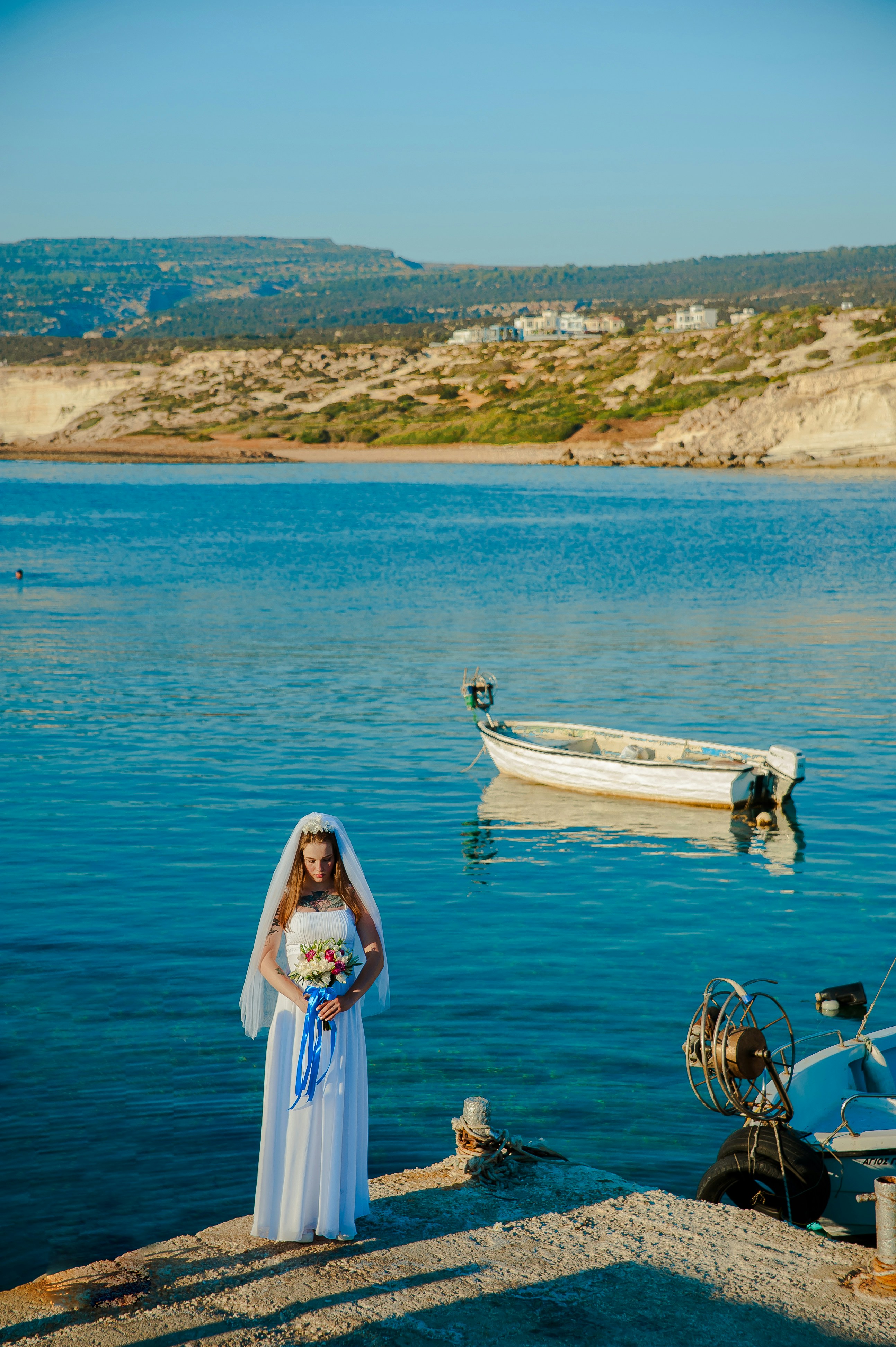 Bride in a flowing white gown holding a bouquet stands on a rocky shore beside a tranquil sea and a small boat. The scene captures the essence of a coastal wedding.