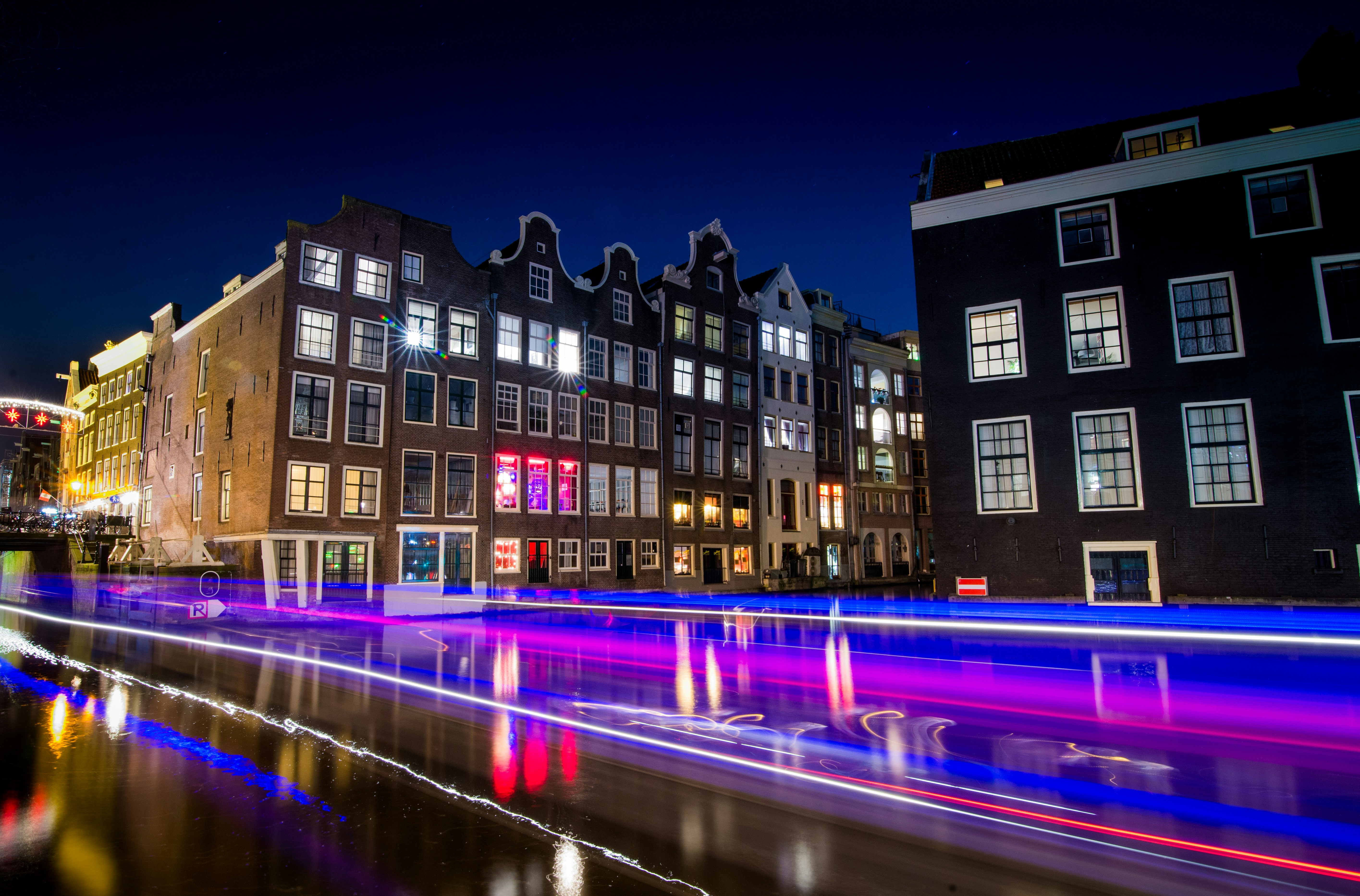 Vibrant light trails from a boat reflect on a canal beside historic buildings under a deep blue night sky.