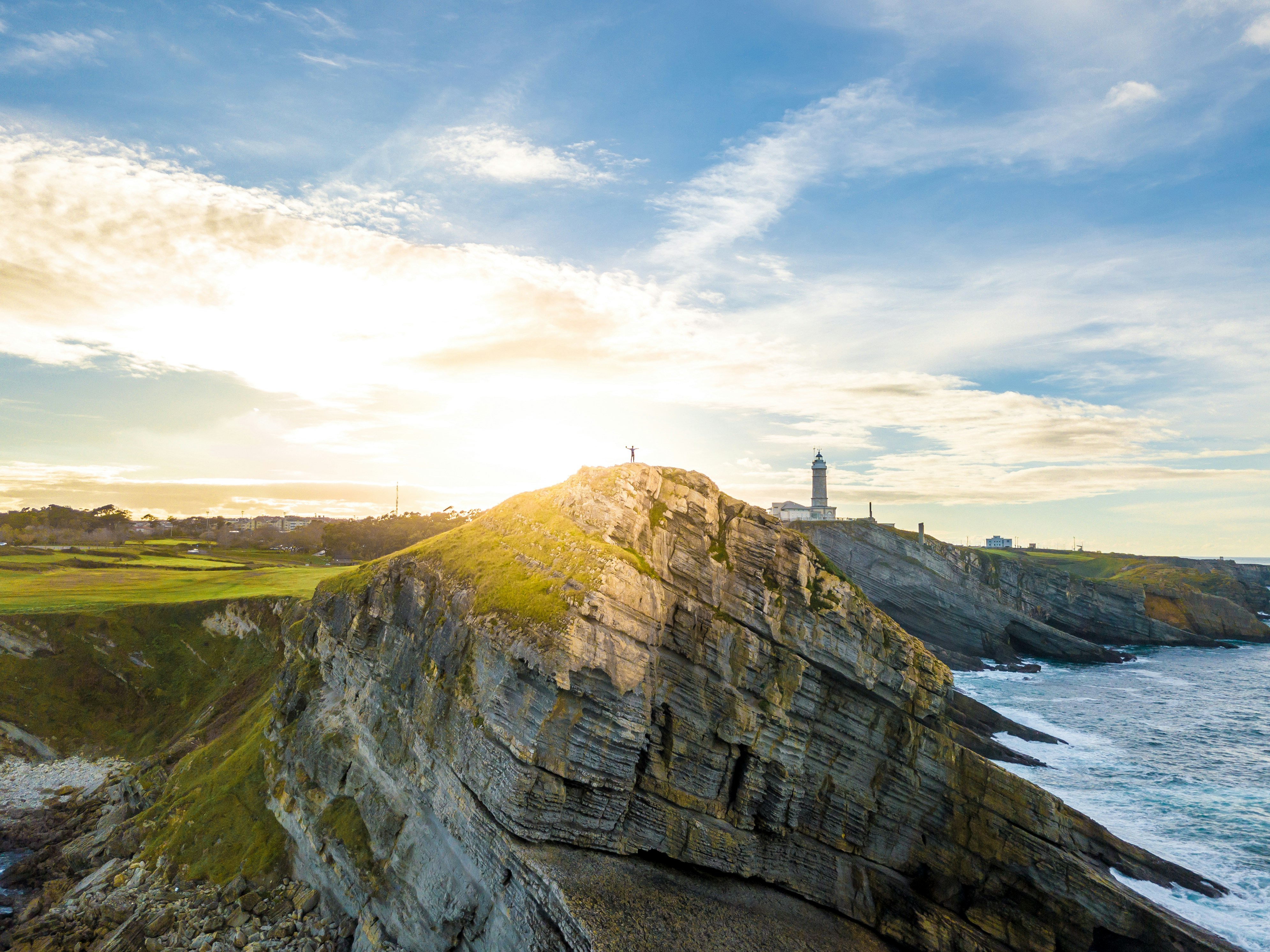 Cliff overlooking the ocean with a lighthouse in the distance under a bright sky.