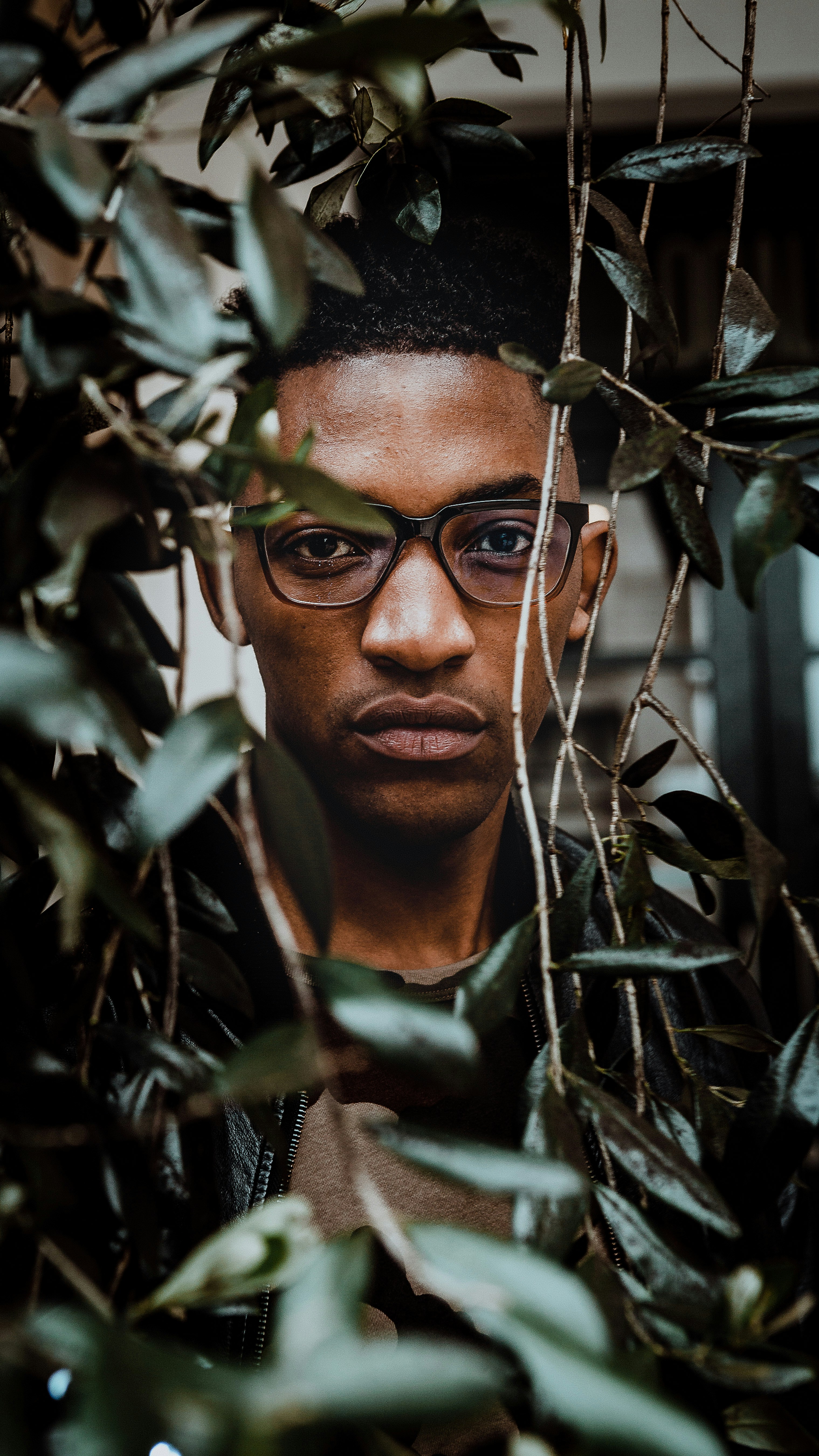 A young man with glasses peers through a tangle of leaves, creating a striking contrast between his expression and the surrounding greenery.