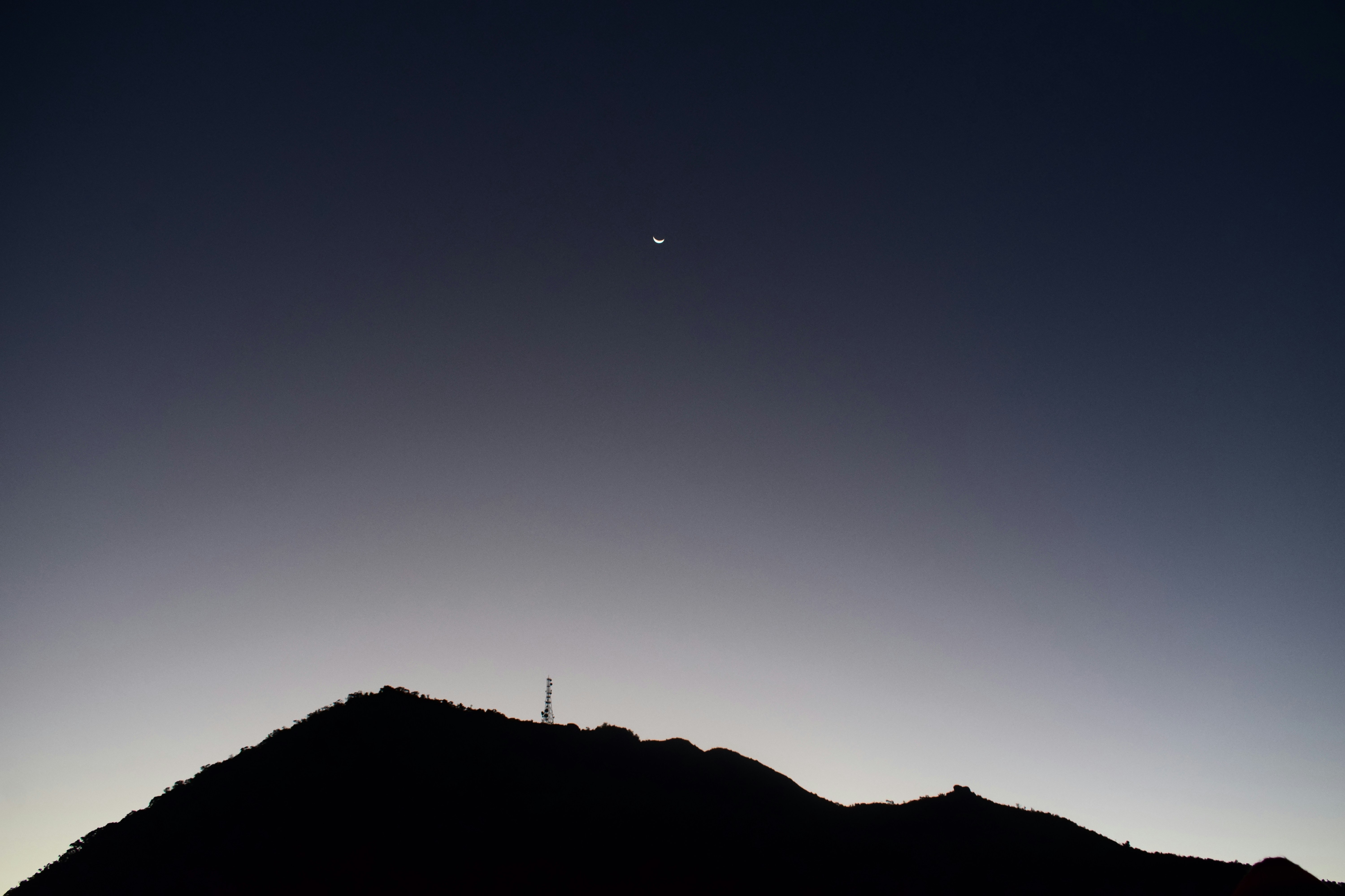 Silhouette of a mountain range under a gradient twilight sky, with a crescent moon peeking above the peaks.