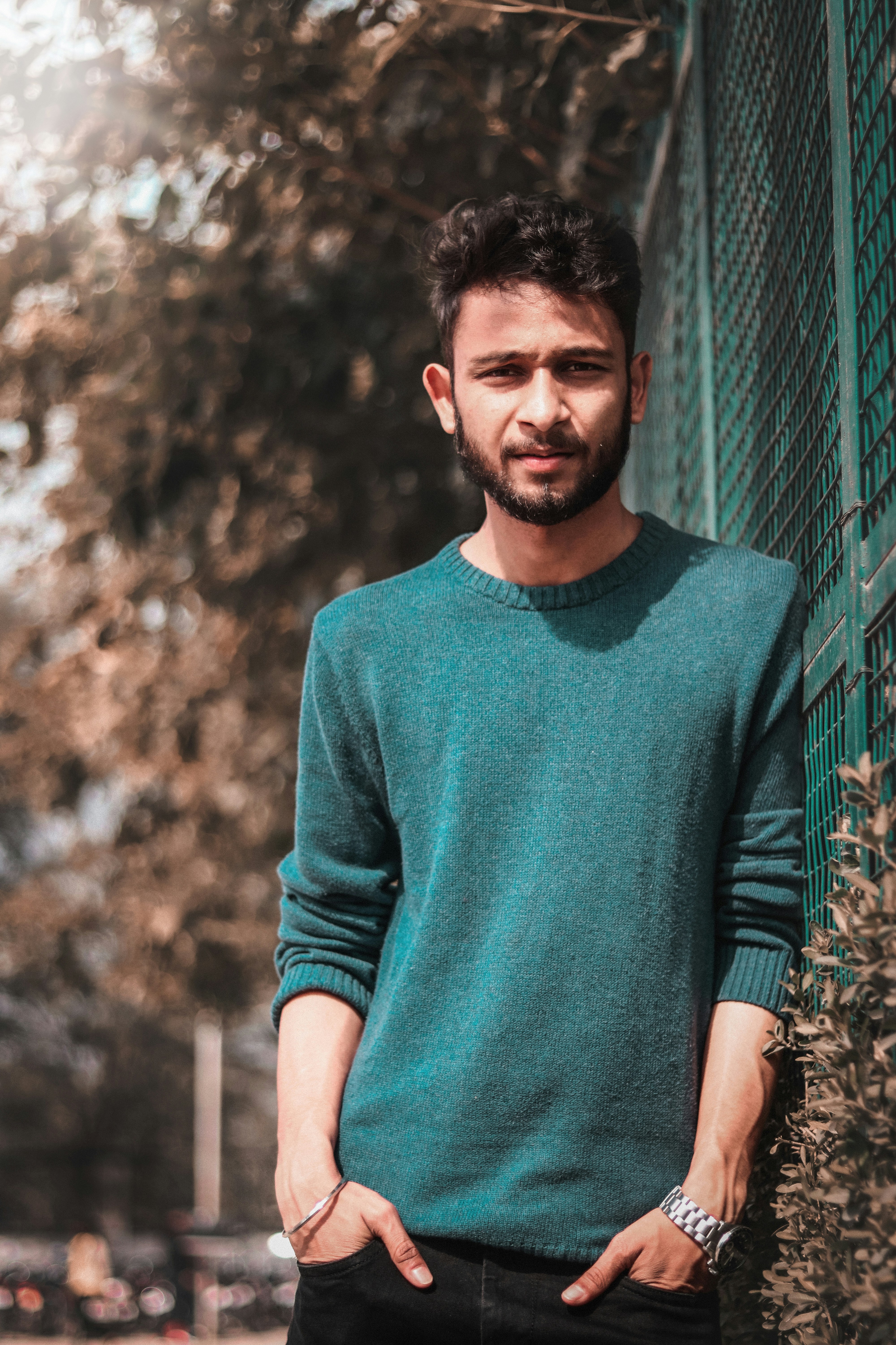 Young man in a teal sweater stands against a green fence, embodying a moment of calm amidst an urban backdrop.