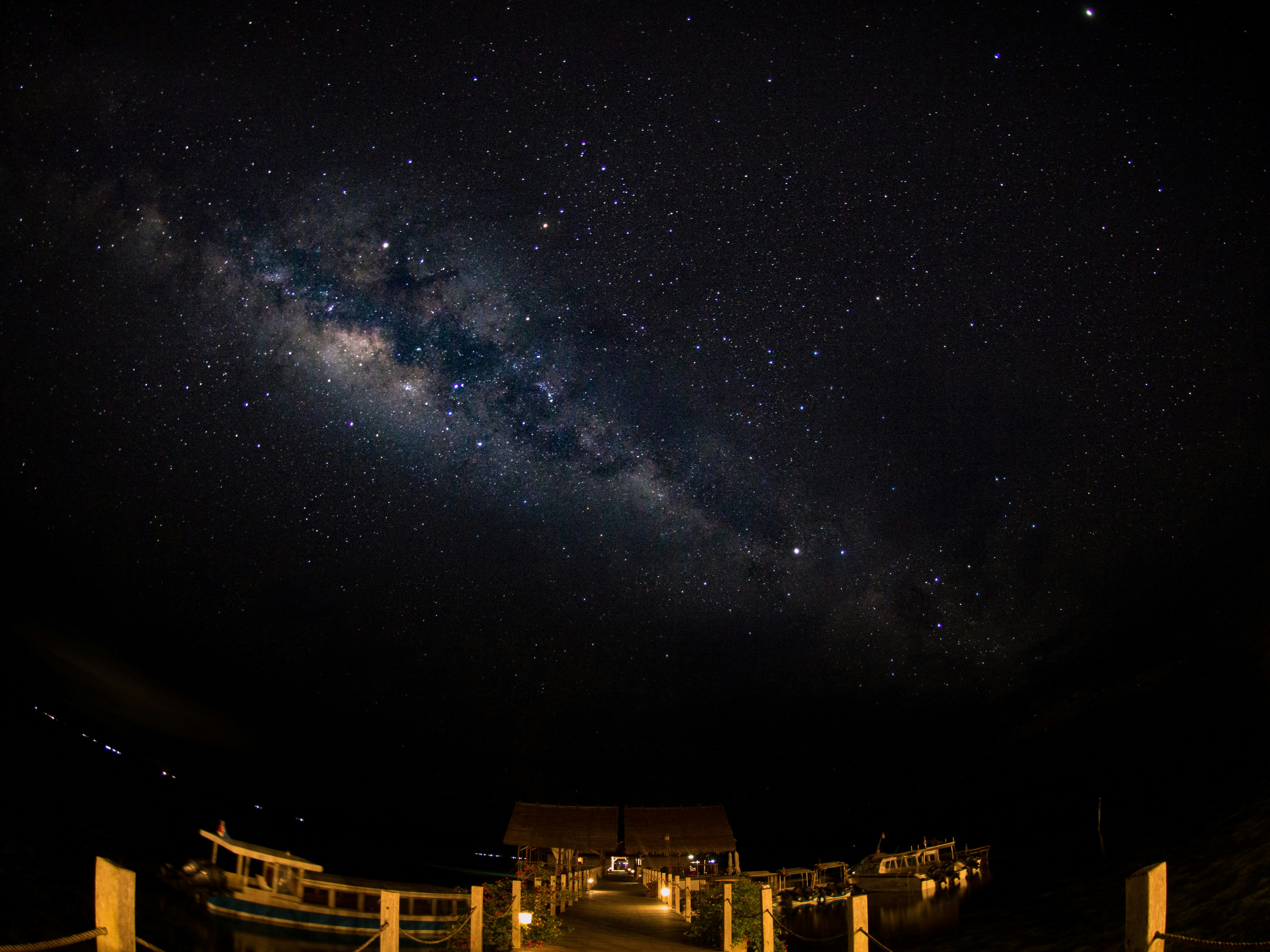 Milky Way arcs across a star-filled night sky above a softly lit wooden pier in a quiet coastal nightscape photograph.