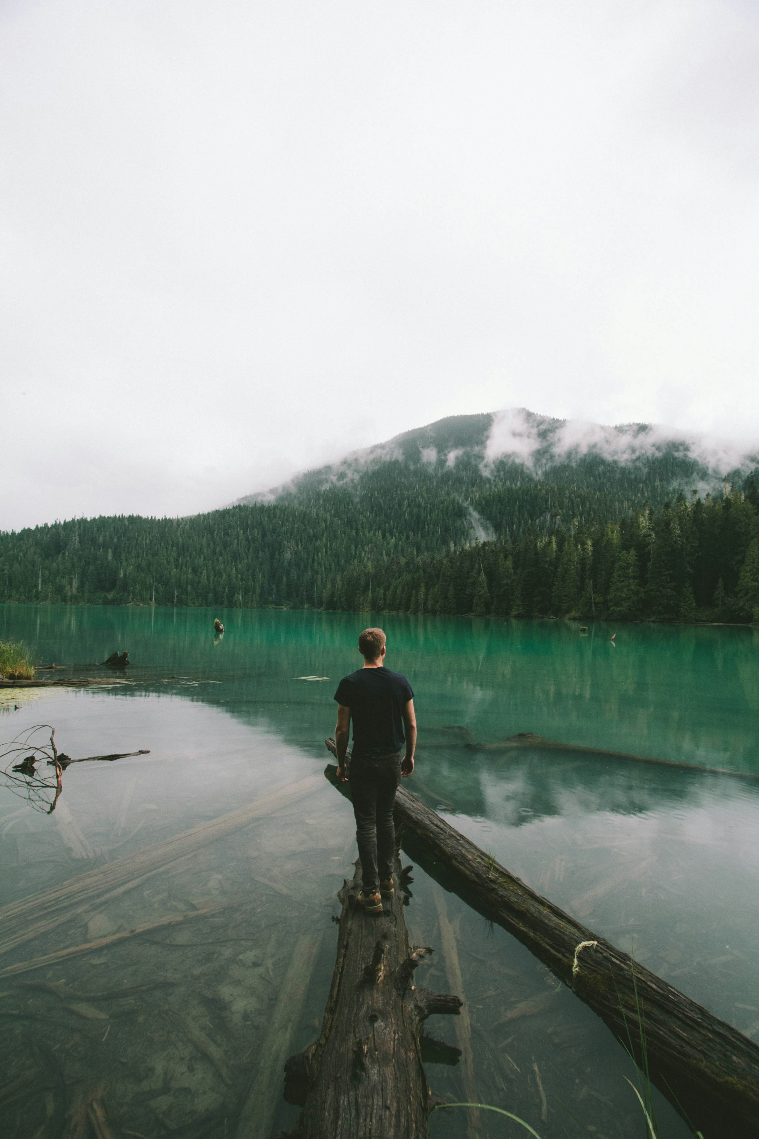A person stands on a log over a serene lake, surrounded by lush green mountains and misty clouds. The still water reflects the vibrant landscape.