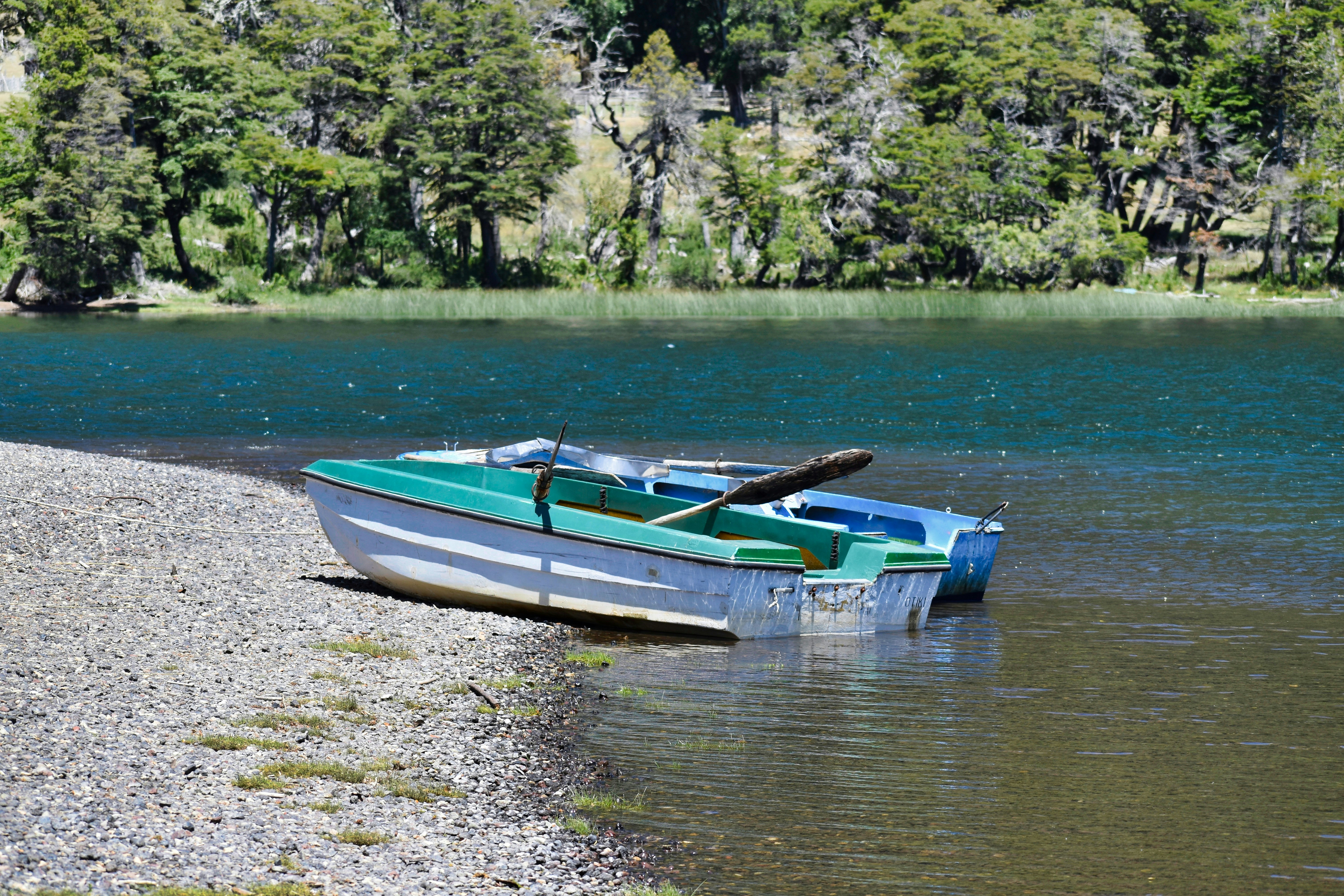 Two boats resting on a pebbly shore beside a tranquil river, surrounded by lush greenery. The scene evokes a sense of peace and natural beauty.