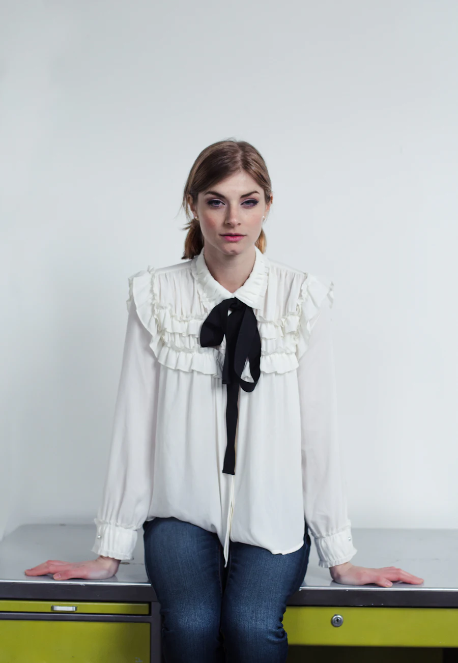 Woman seated against a plain light background, wearing a white ruffled blouse with a black ribbon tie and jeans. Her hands rest on a table edge, creating a simple studio portrait.