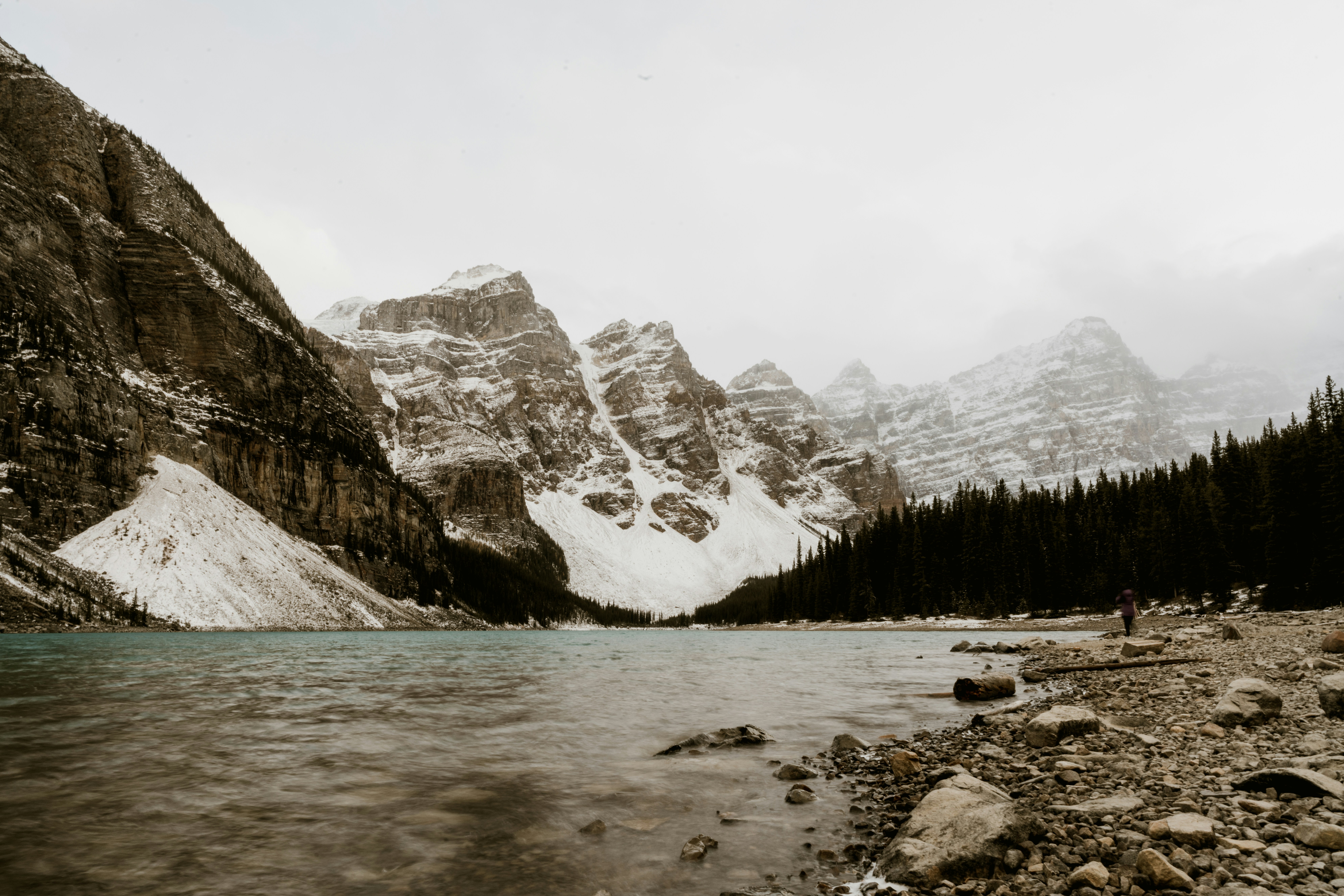 Snow-capped mountains rise majestically above a tranquil lake, framed by a rocky shoreline and dense coniferous forest.