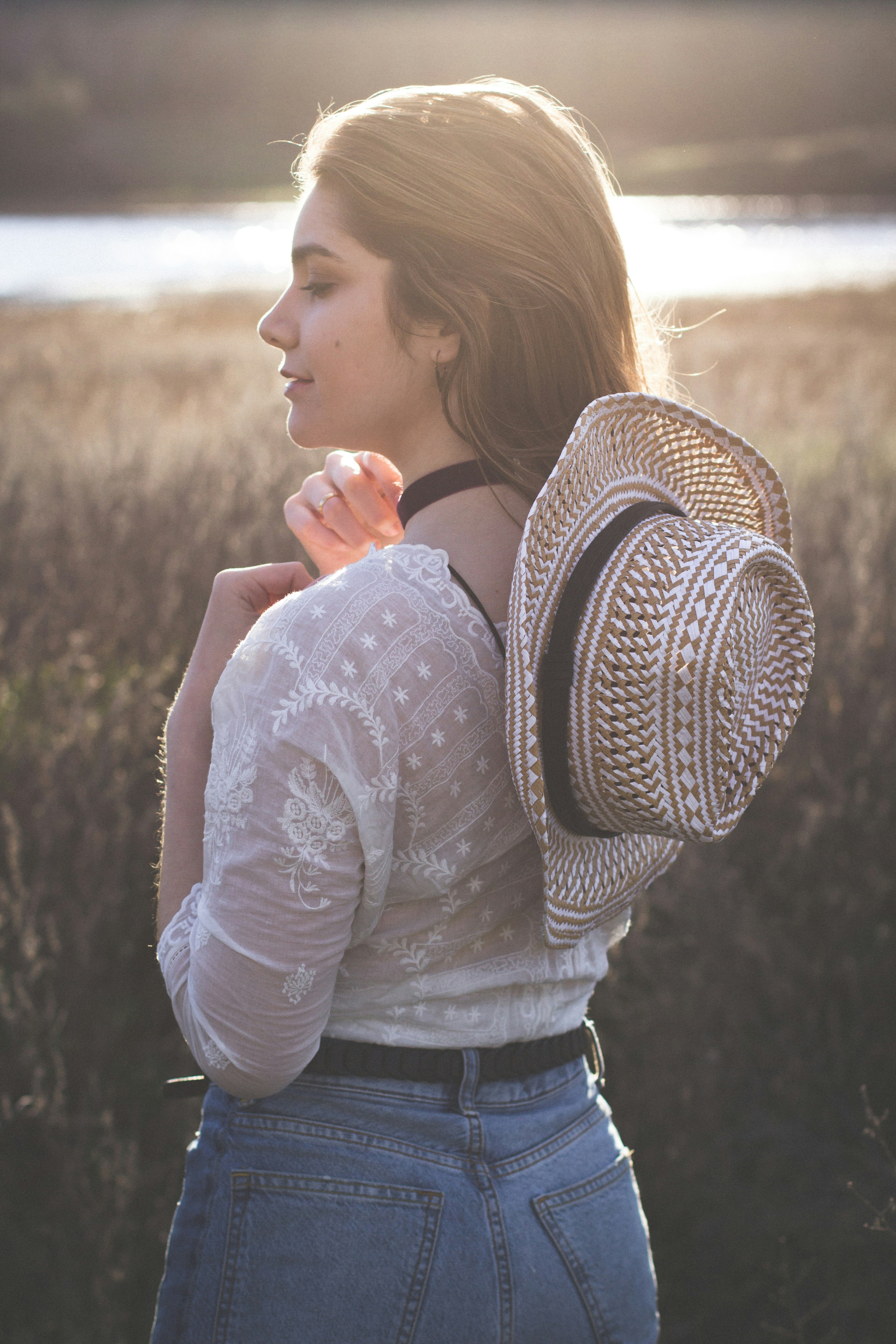 Young woman with flowing hair, gently holding a straw hat against her back, stands amidst golden grass by a shimmering lake.