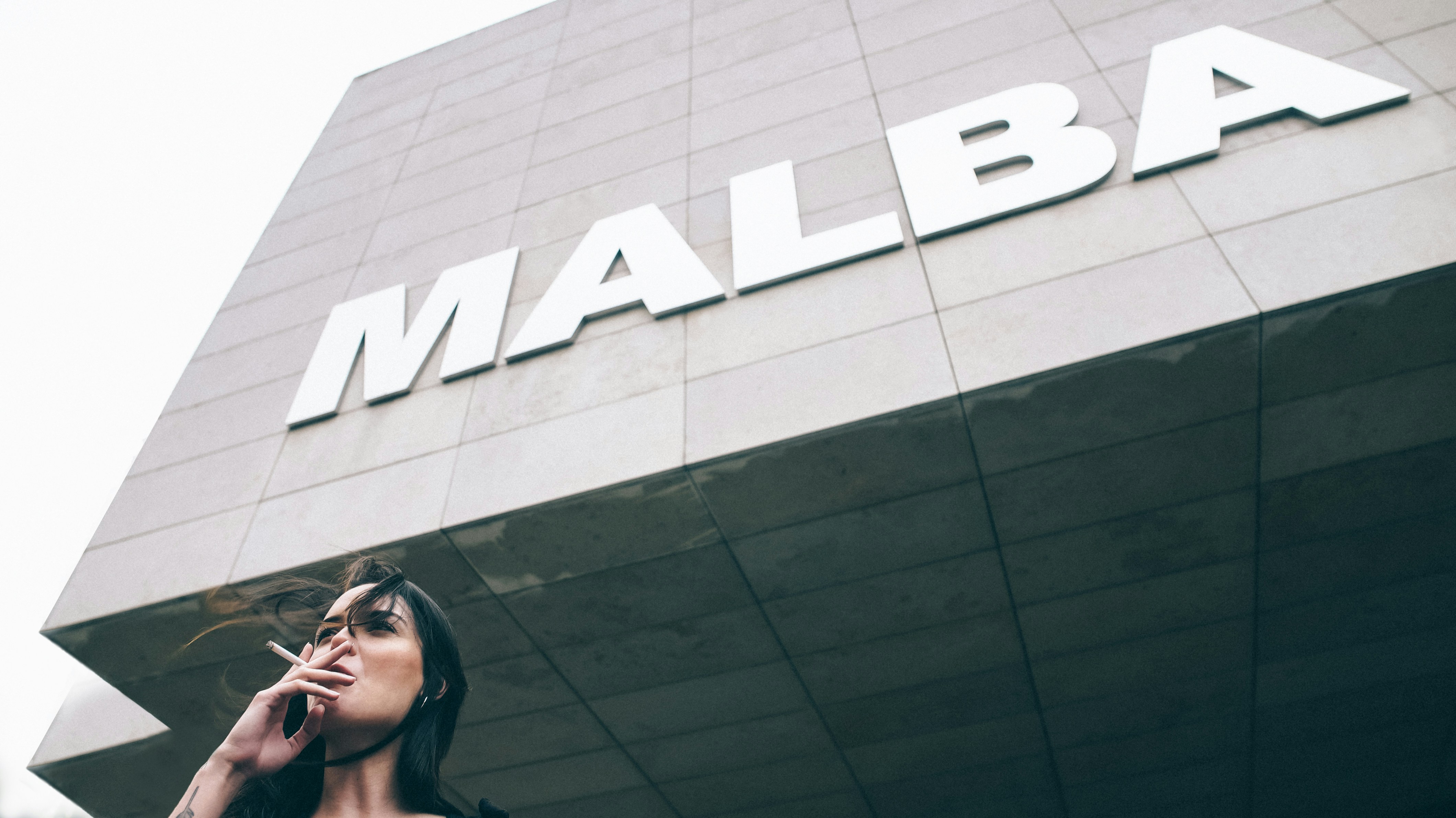 A woman stands outside the MALBA museum, inhaling smoke against a backdrop of modern architecture. The bold letters of 'MALBA' loom overhead.