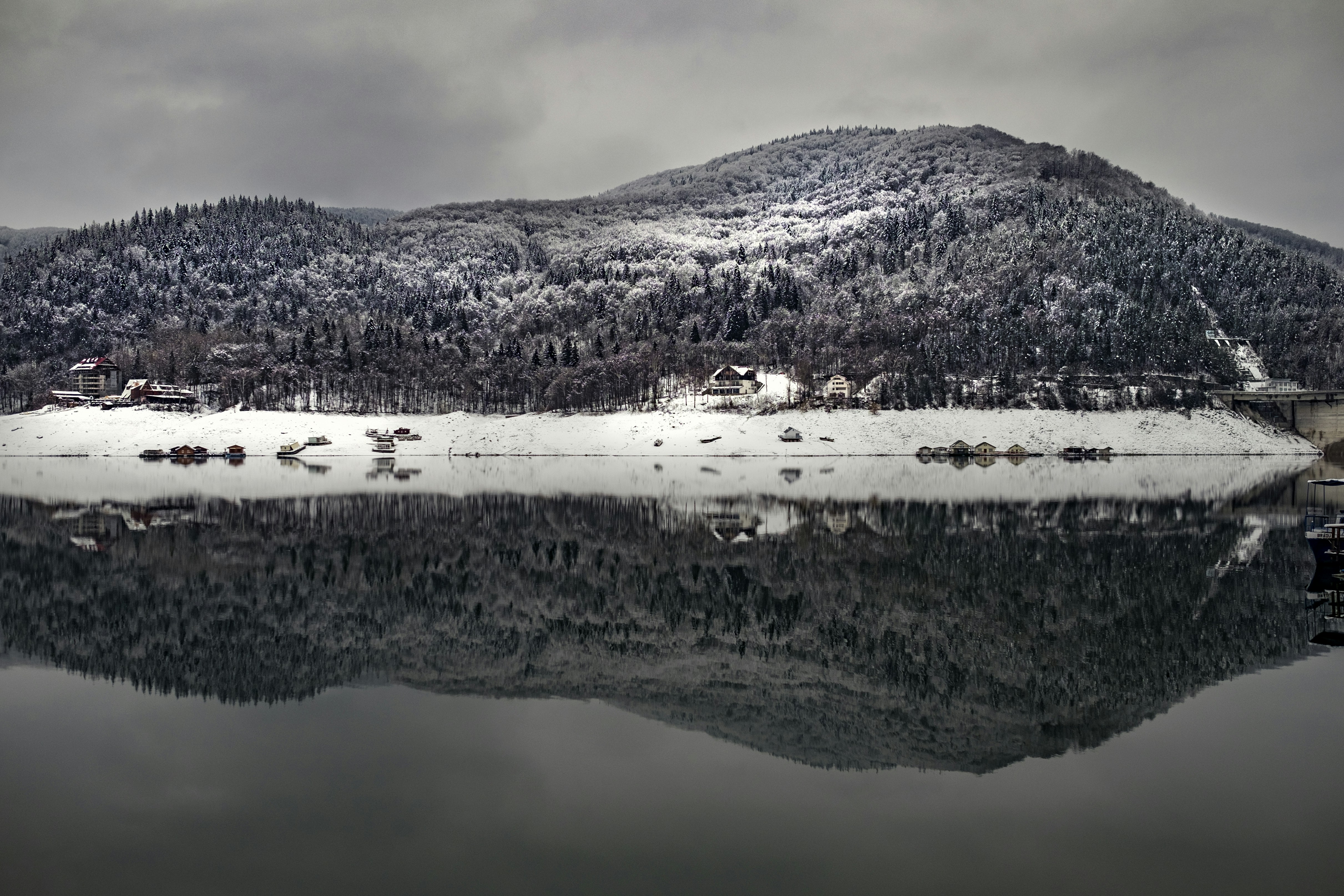 Snow-covered mountains and trees mirrored in a still lake under a cloudy sky.