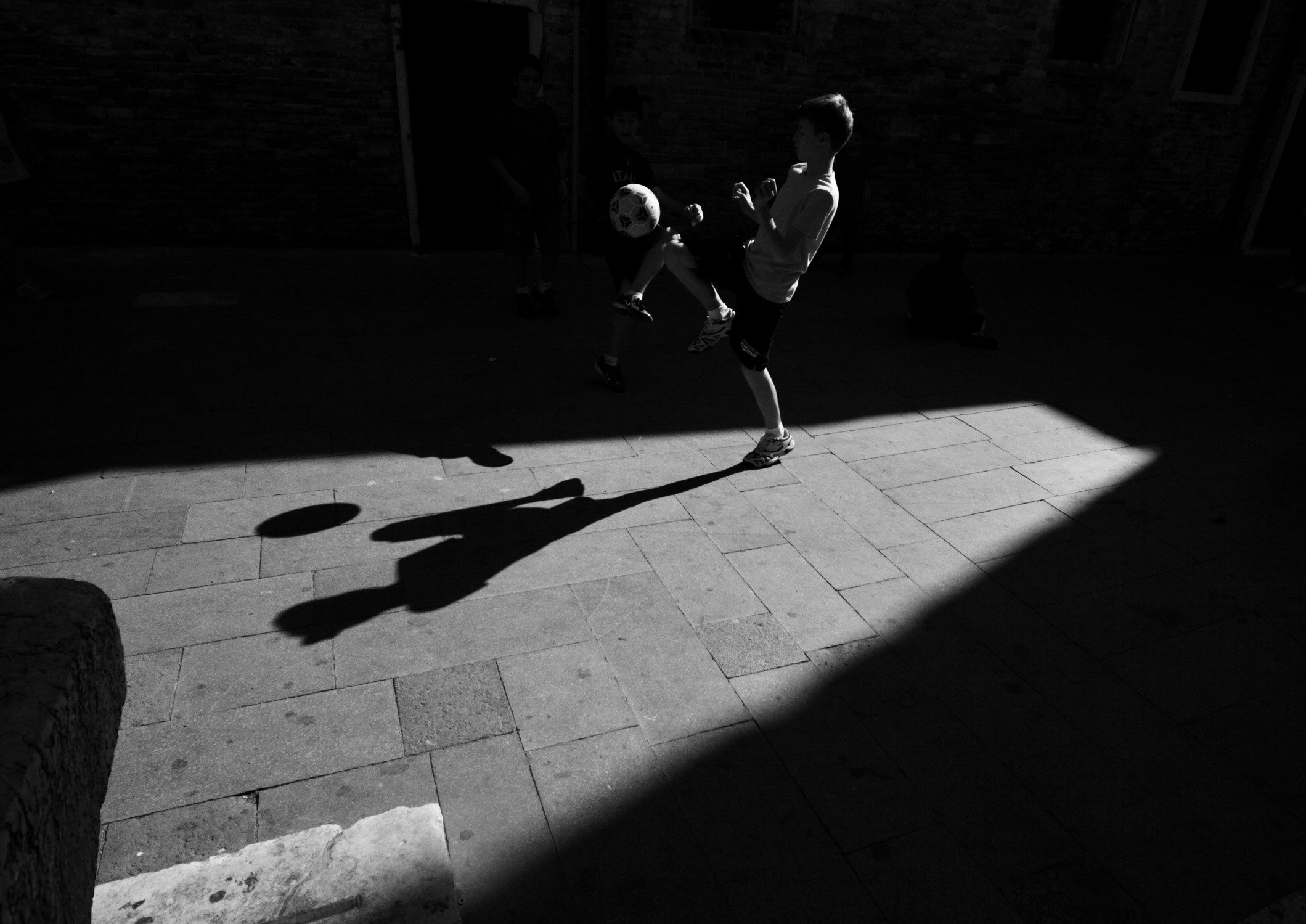 A young boy skillfully juggling a soccer ball in a sunlit courtyard, with dramatic shadows enhancing the scene's dynamic energy.