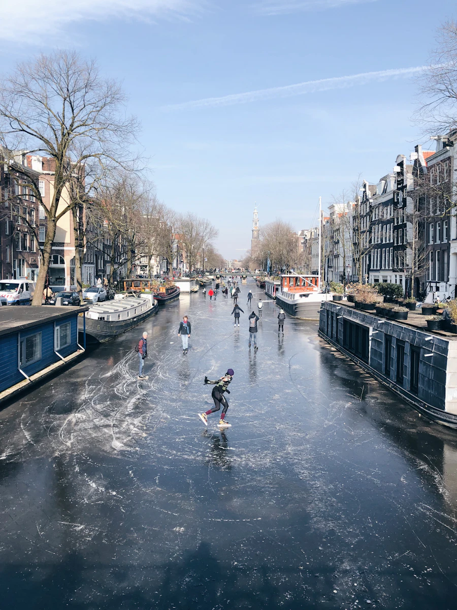 A lone skater glides across a sunlit, glassy canal lined with historic townhouses and moored boats. The distant cityscape recedes toward a tower as others skate along the ice.