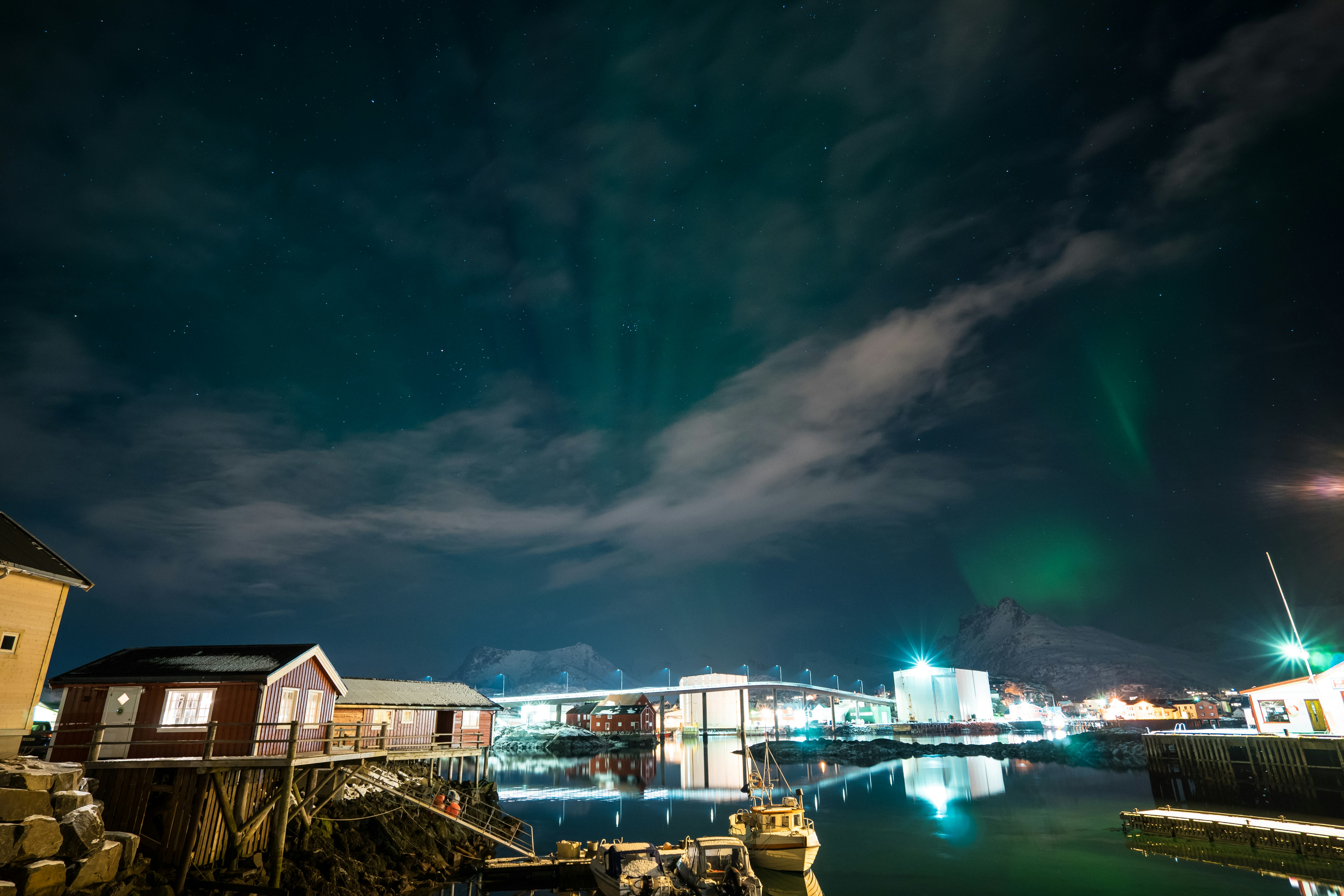 Northern lights dance above a serene harbor, with quaint wooden houses lining the shore. The reflection in the water enhances the ethereal atmosphere.