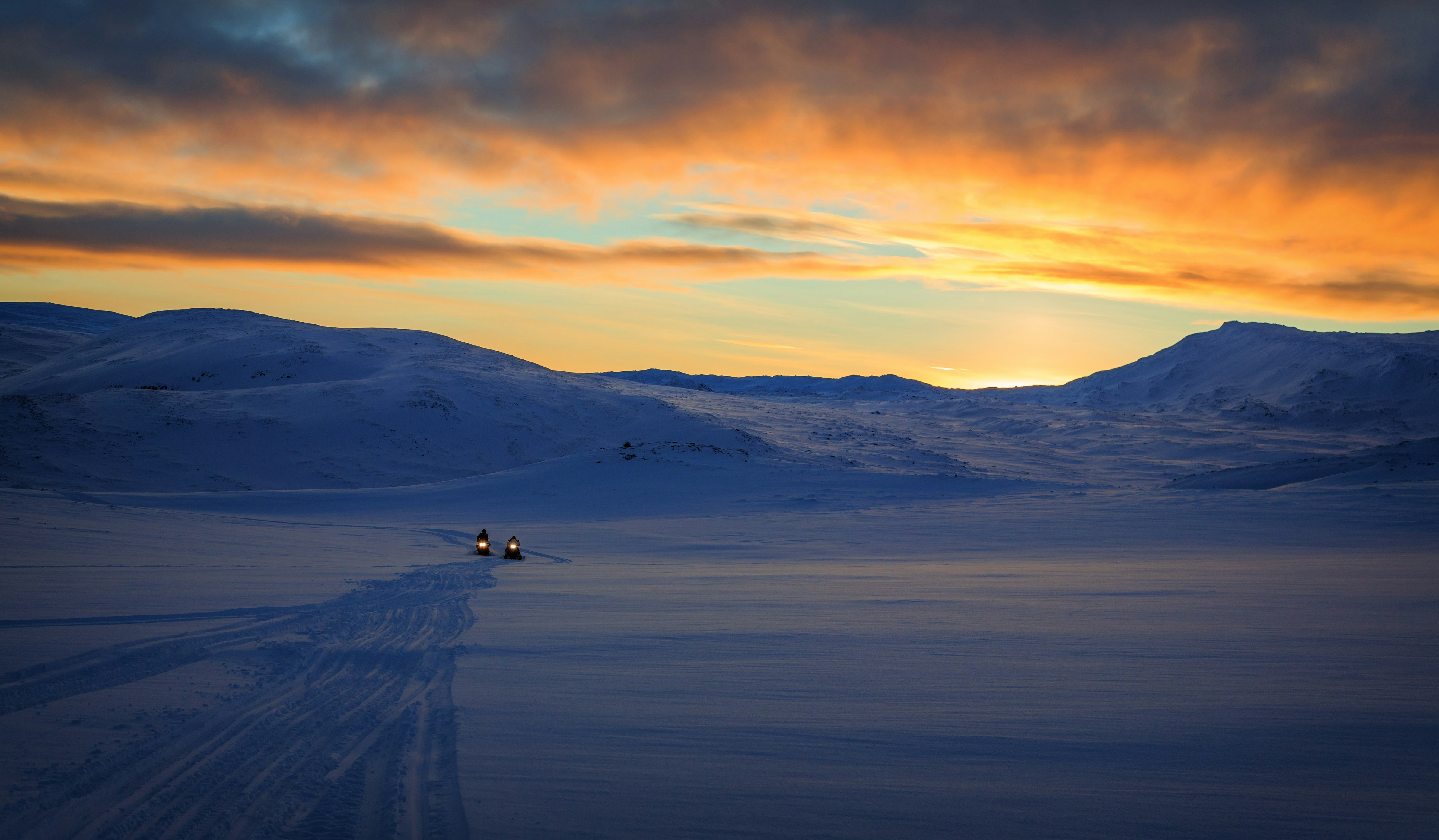 Two snowmobiles traverse a vast, snowy landscape under a vibrant sunset, highlighting the serene beauty of winter's embrace.