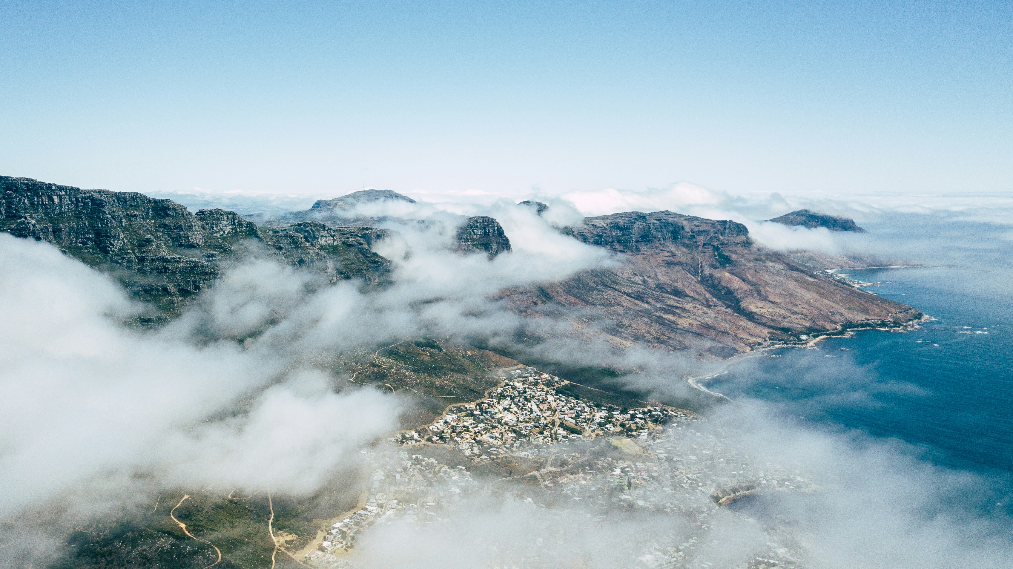 Aerial view of a coastal town nestled between majestic mountains and a blanket of clouds, capturing the harmony of nature and human settlement.