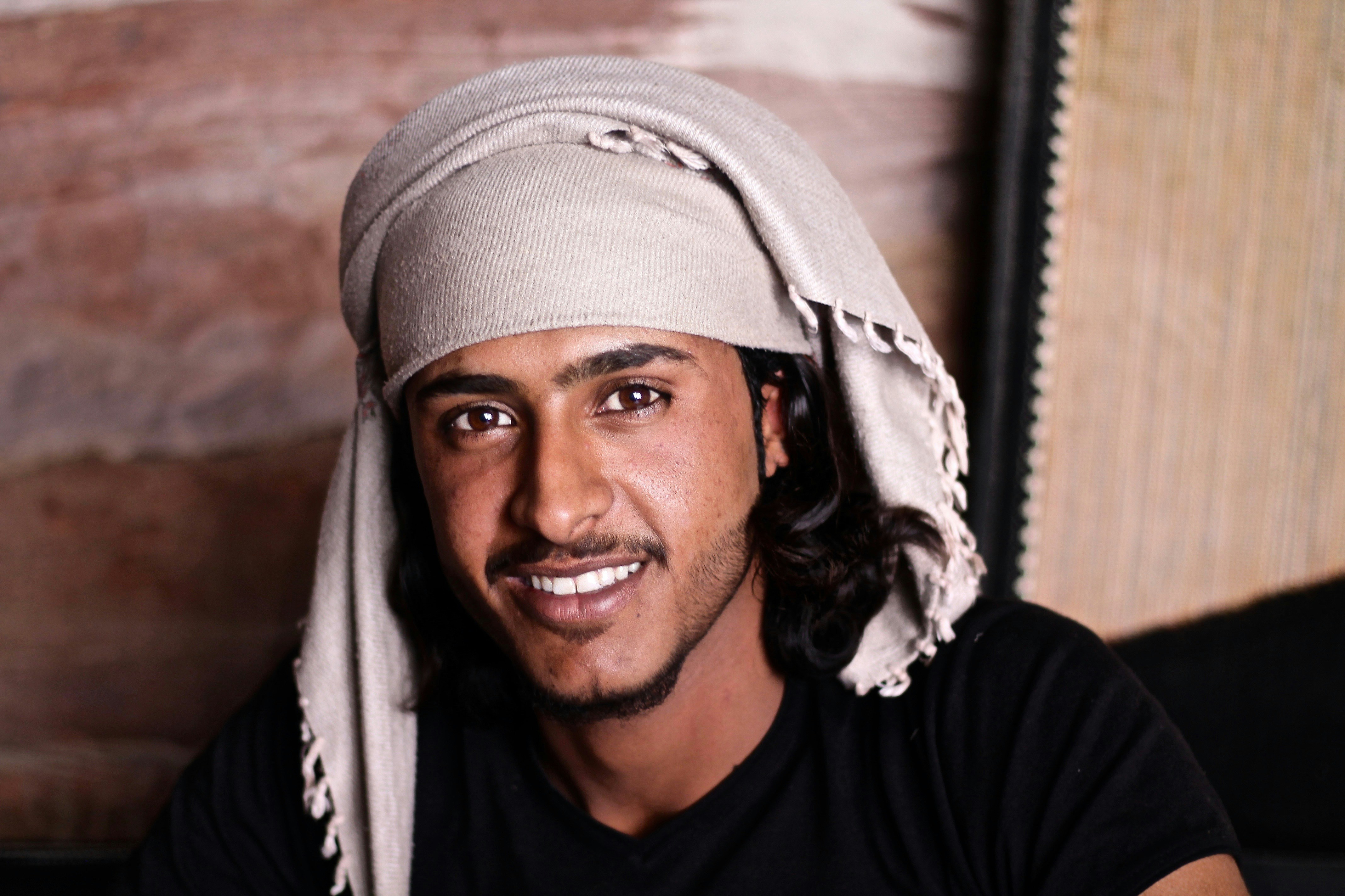 This portrait captures a man with a warm smile, draped in a light beige turban against a textured, earthy backdrop. The soft lighting accentuates his expressive eyes and contrasts beautifully with his dark hair and black shirt, adding depth to the image. The harmonious blend of natural tones and the subject's inviting expression create a welcoming and engaging atmosphere.