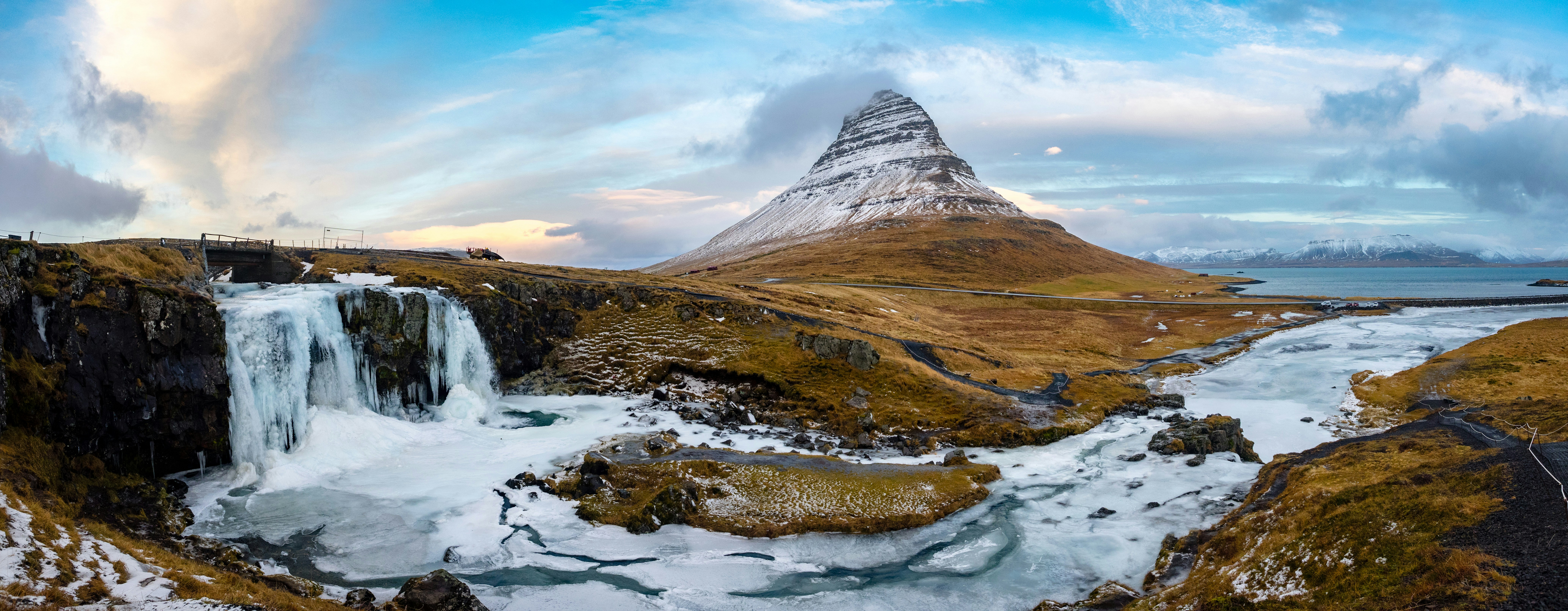 Icy waterfall cascading into a river with Kirkjufell mountain in the background, under a dramatic sky. The scene captures the tranquility of winter in Iceland.