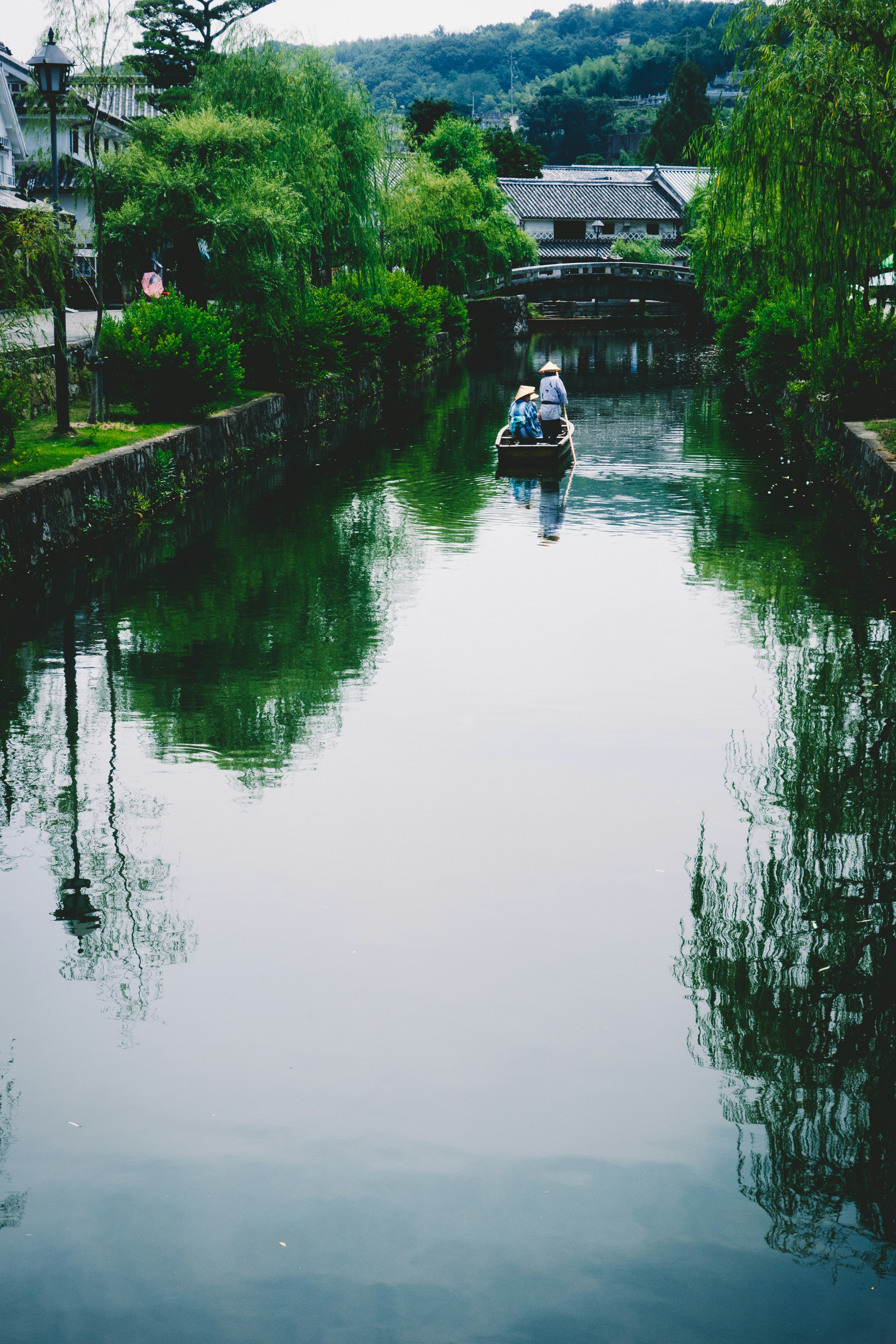 Two individuals in a small boat glide peacefully along a tranquil canal, surrounded by lush greenery and reflections in the water.