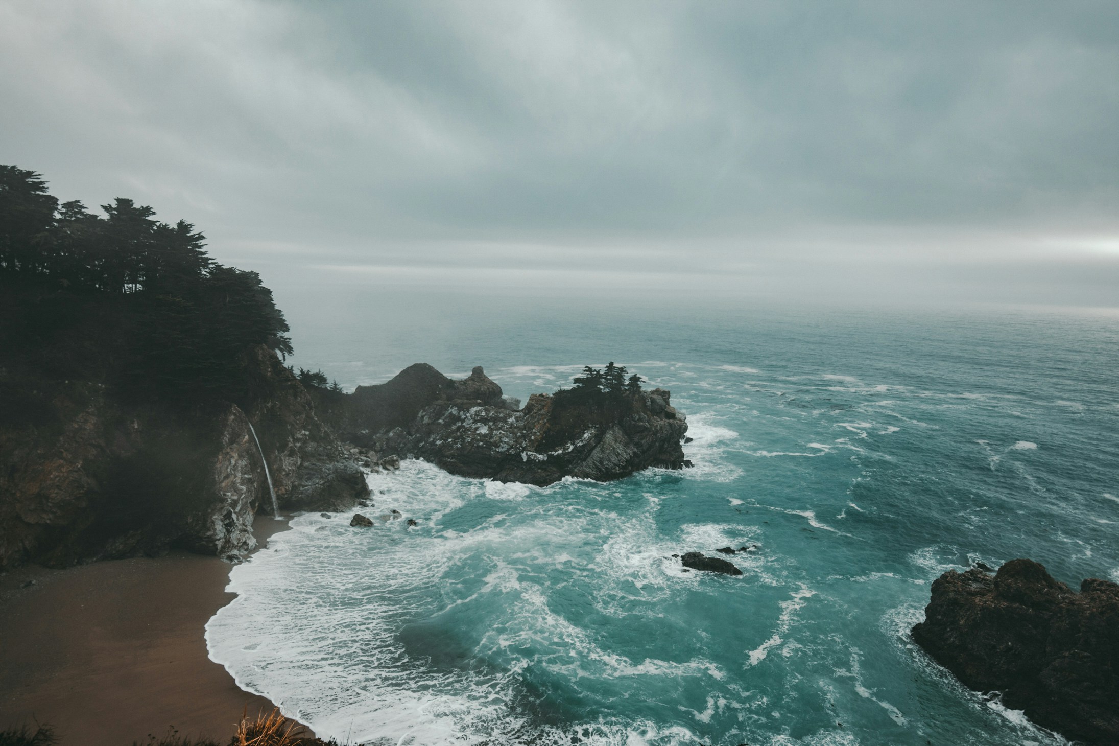 This captivating image captures the rugged beauty of a coastal landscape, with dramatic cliffs enveloped by misty clouds. The ocean's turquoise waves crash against the rocks, creating a dynamic interplay of motion and stillness. The muted color palette, dominated by soft blues and grays, enhances the tranquil yet mysterious atmosphere, making the scene both visually striking and deeply immersive.